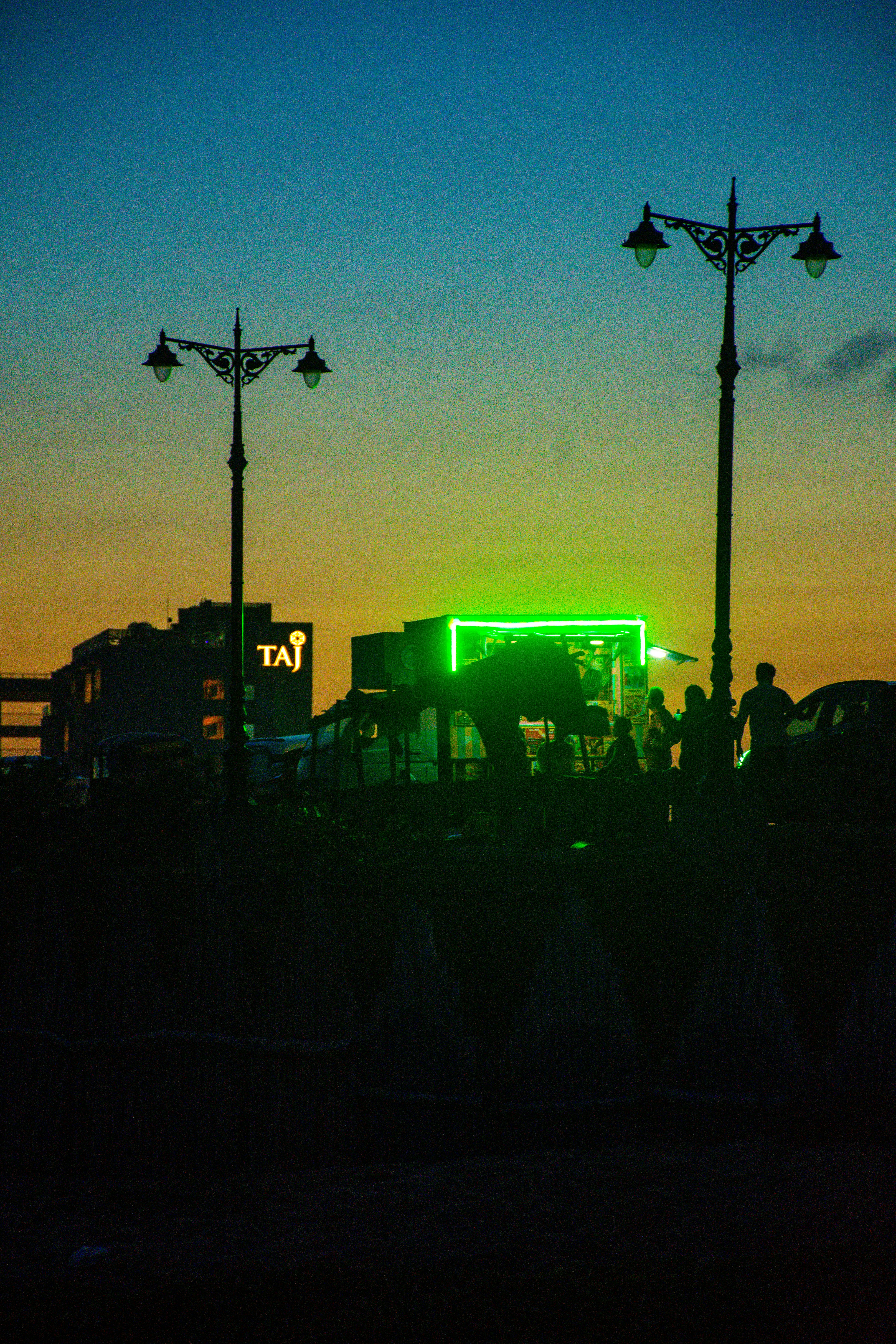 Silhouetted figures gather around a neon-lit food cart under street lamps as the sun sets, creating a vibrant contrast against the twilight sky.