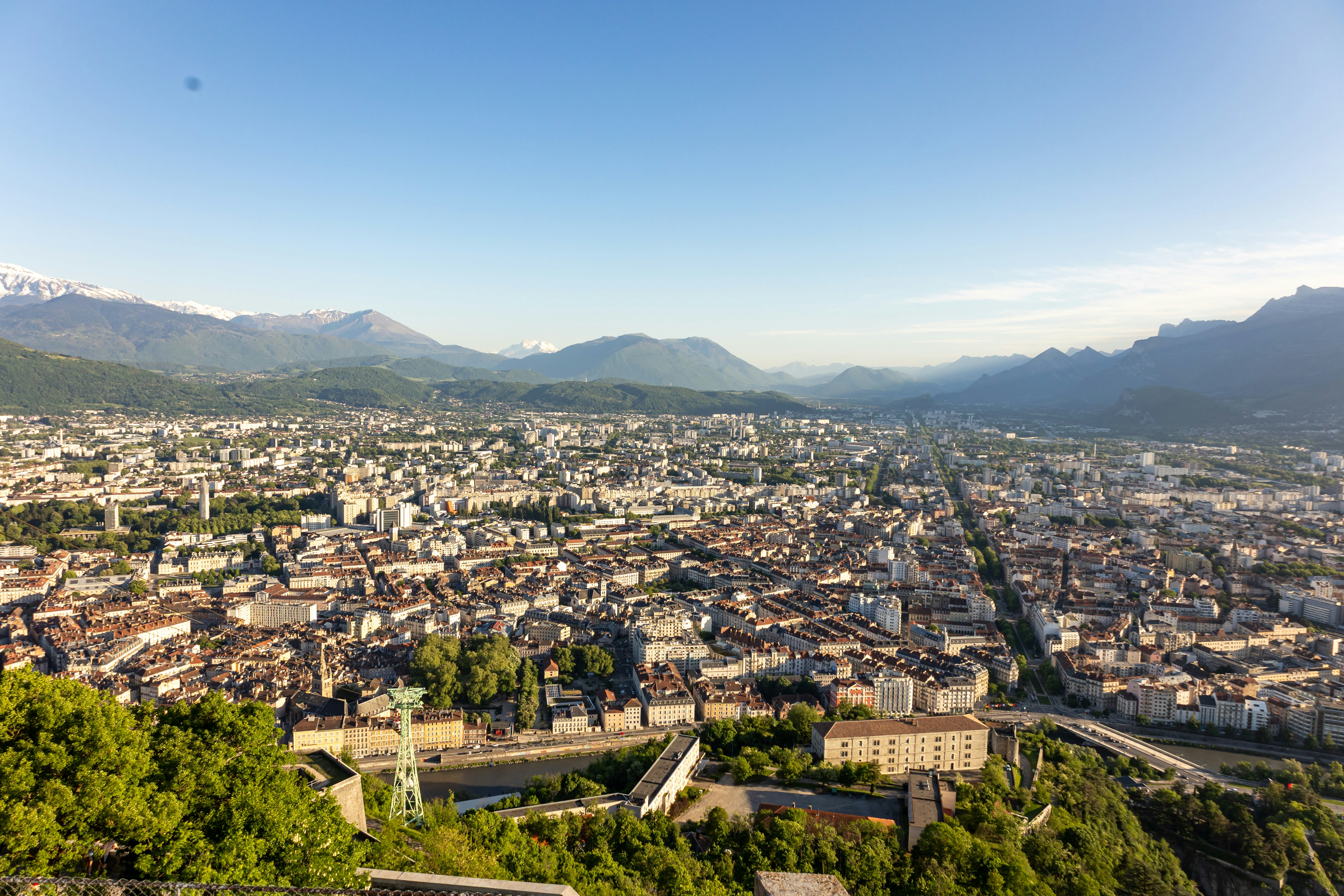 Vast aerial view of a city nestled between mountains, showcasing a blend of greenery and urban architecture. The landscape highlights the interplay of natural and built environments.