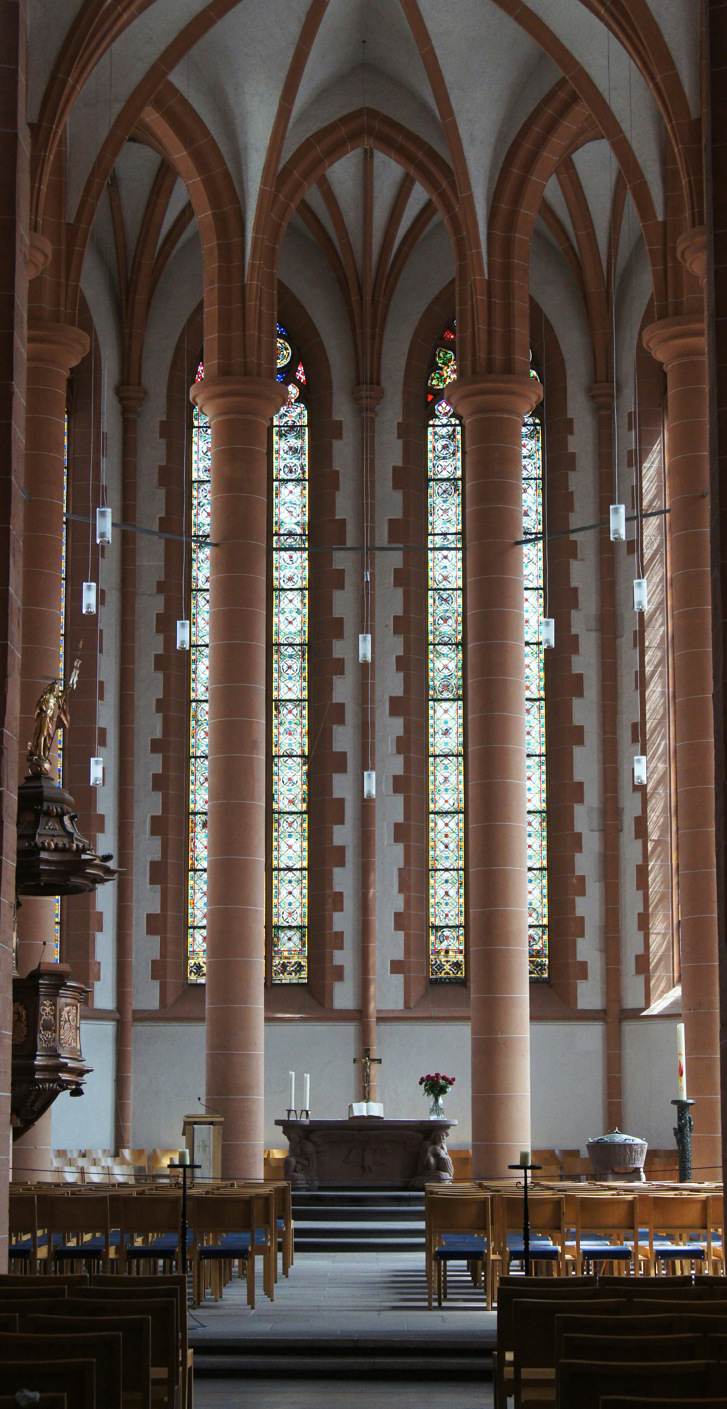 Interior view of a church showcasing tall arches and stained glass windows, with a central altar and wooden pews arranged in a serene setting.