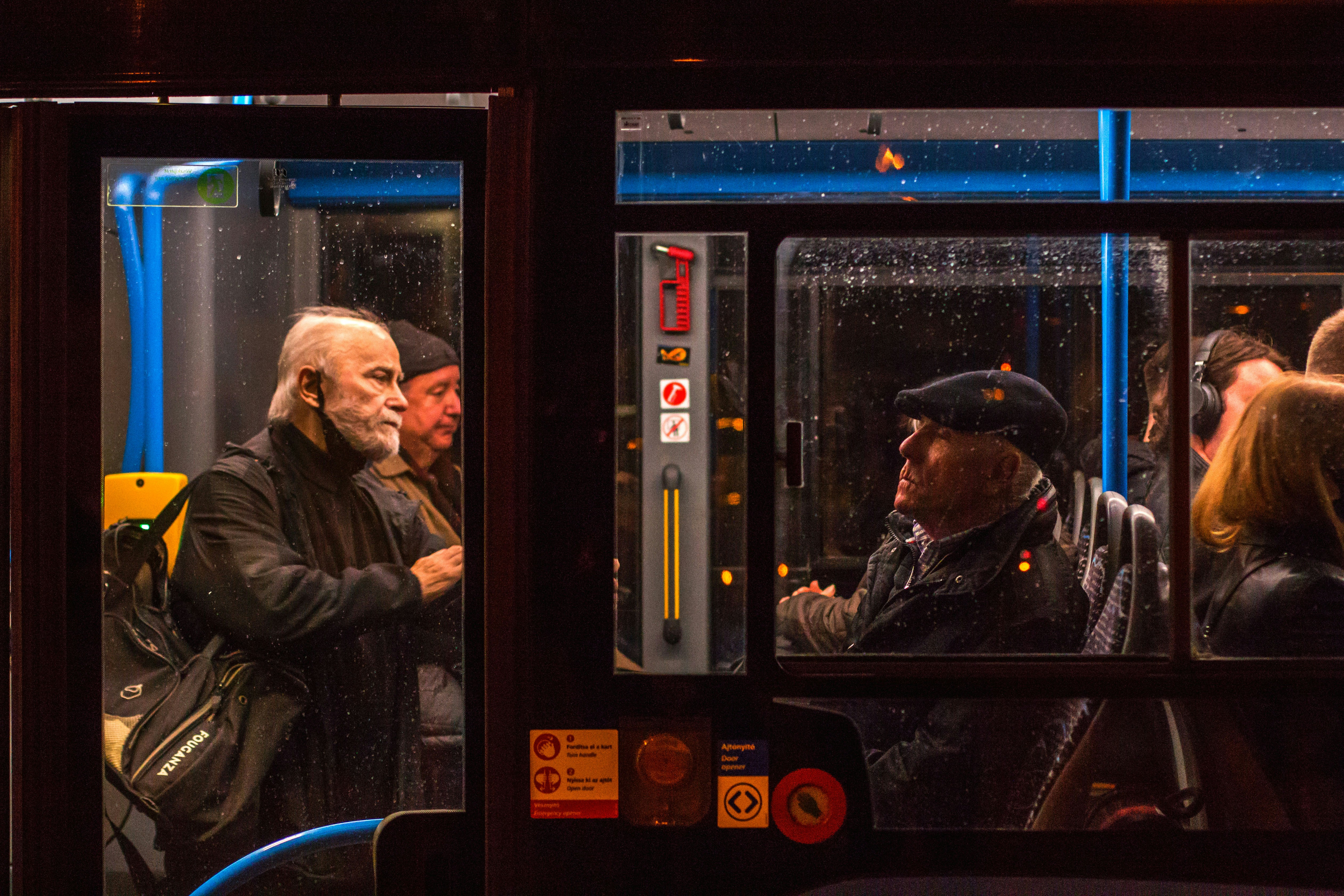 Evening Wait – Elderly Gentlemen Heading Home in the Rain" Three elderly men stand under the soft glow of a bus stop light, their faces etched with patience as they wait for the evening bus. Raindrops cling to the windows of nearby vehicles, blurring the city lights into streaks of gold and silver. Their postures—perhaps leaning on canes or clutching worn briefcases—hint at lifetimes of routine. The scene is a quiet ode to twilight rituals, companionship, and the dignity of small journeys. | People ride a bus at night, illuminated by lights.