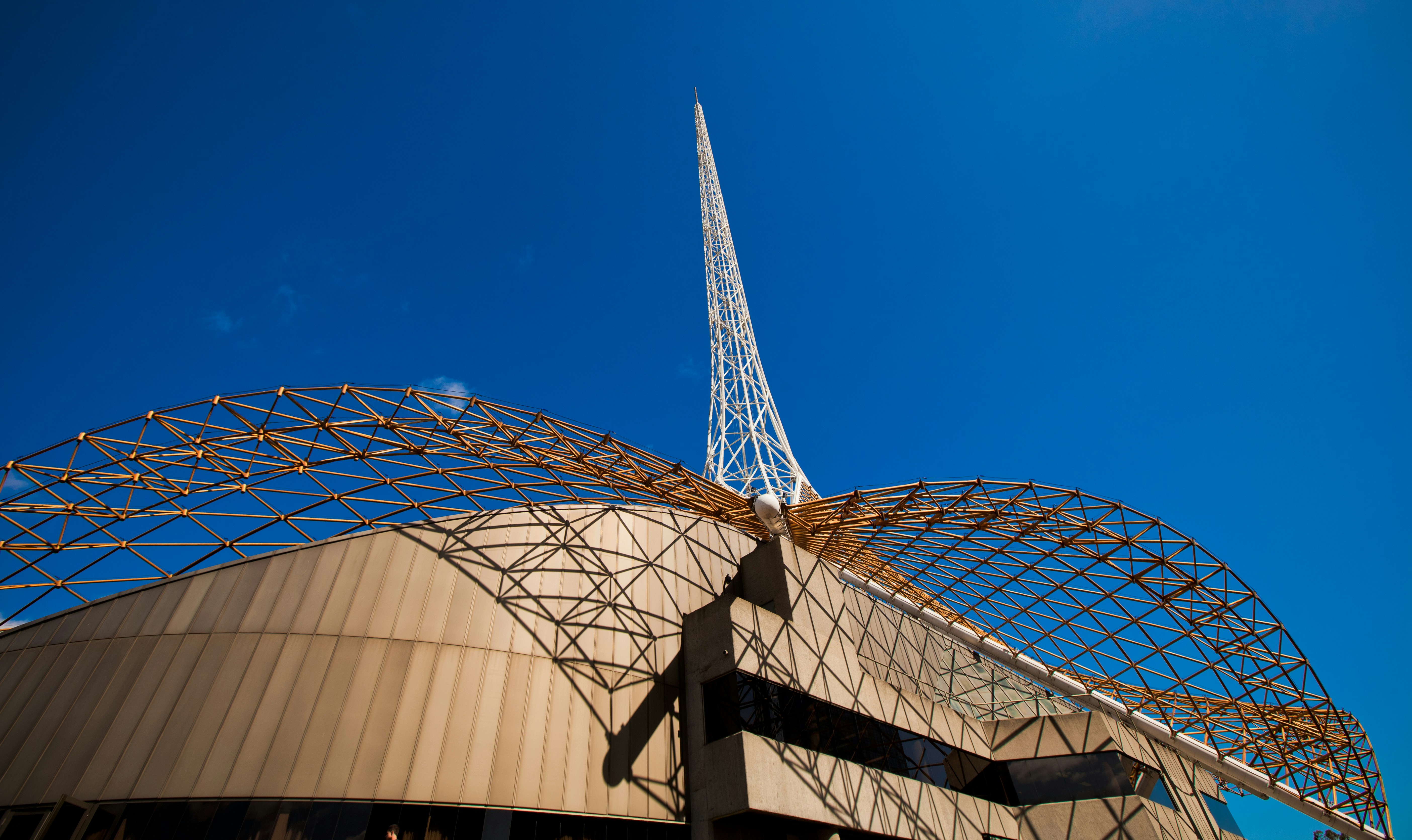 A striking shot of the Arts Centre Melbourne, featuring its unique curved roof and towering spire set against a bold blue sky. A modern architectural icon in the cultural heart of the city. | A modern building pierces the sky.