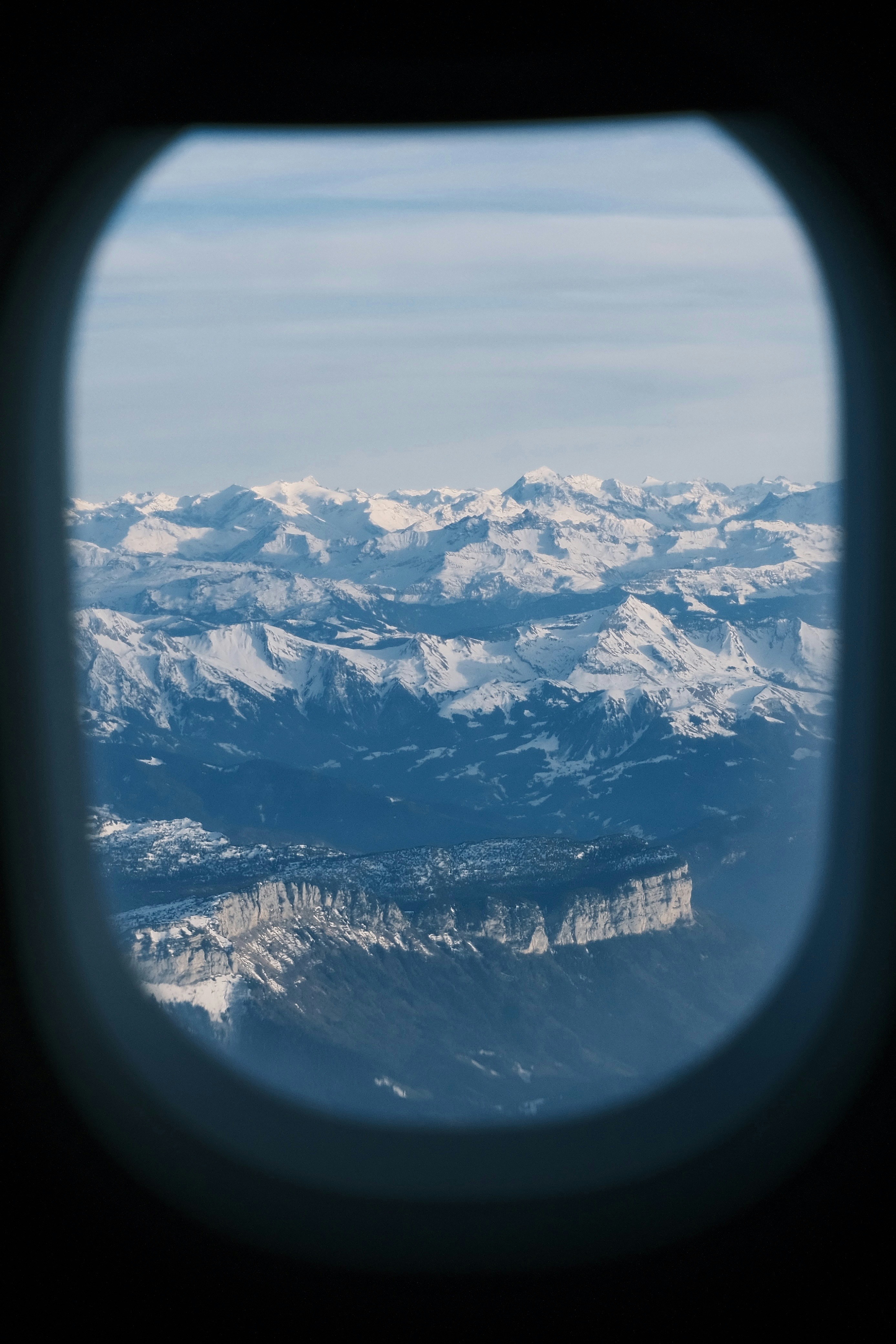 Snow-capped mountains viewed through an airplane window, showcasing rugged terrain and a clear sky. The scene captures the essence of travel and adventure.