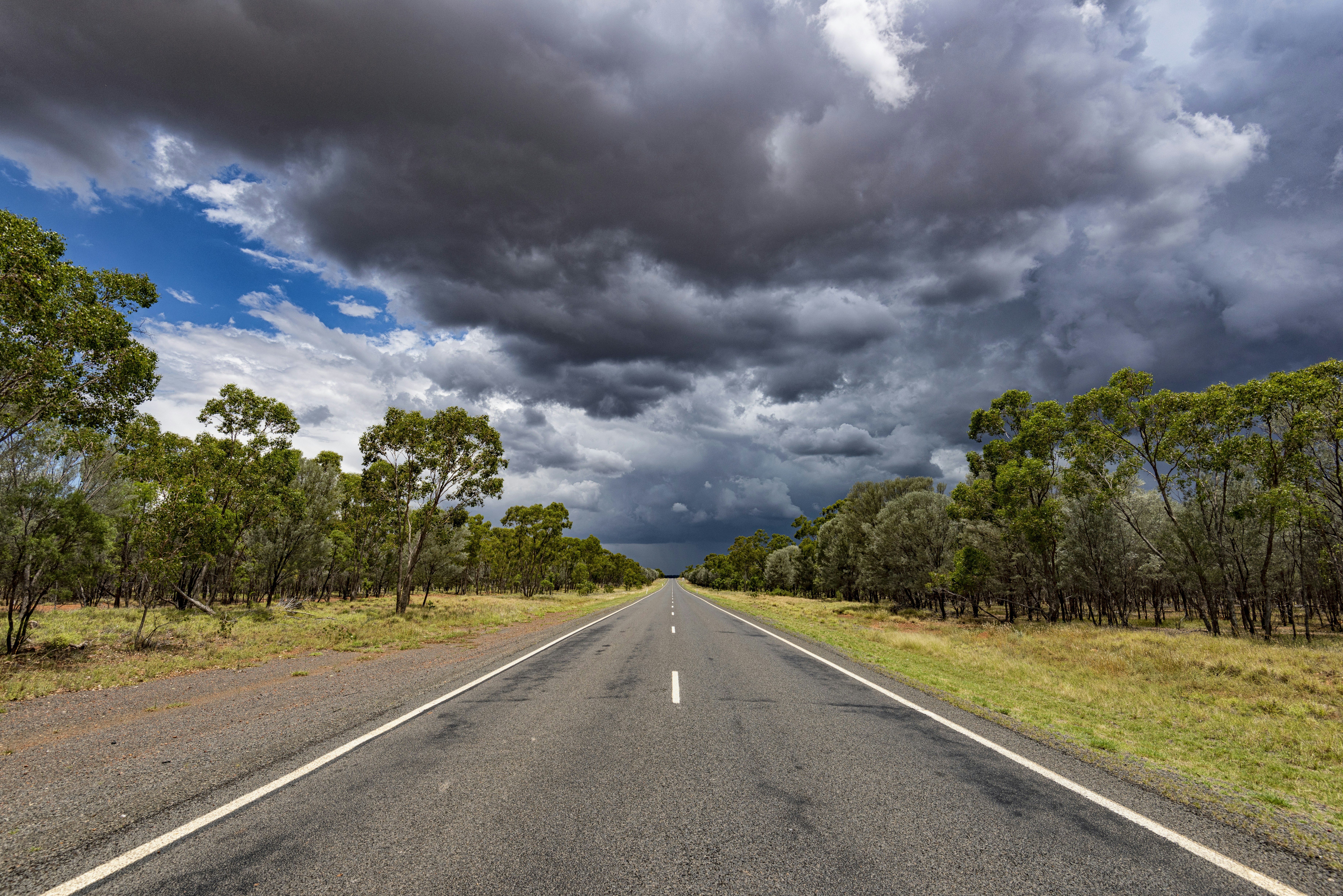 A long, straight road stretches through a landscape flanked by trees under a dramatic sky filled with dark clouds. The scene evokes a sense of adventure and anticipation.