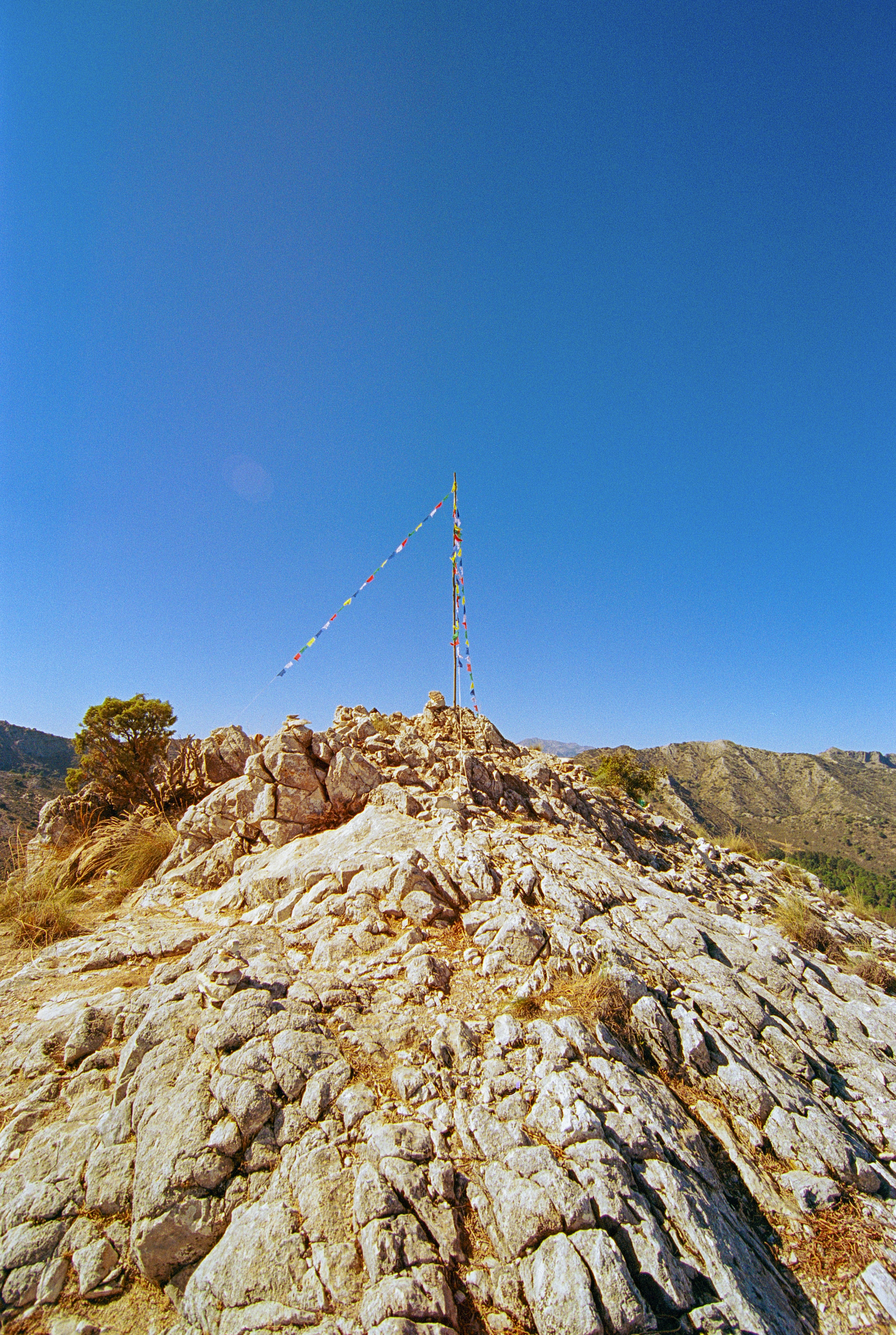 Colorful prayer flags flutter atop a rocky summit, surrounded by expansive mountain views under a clear blue sky.