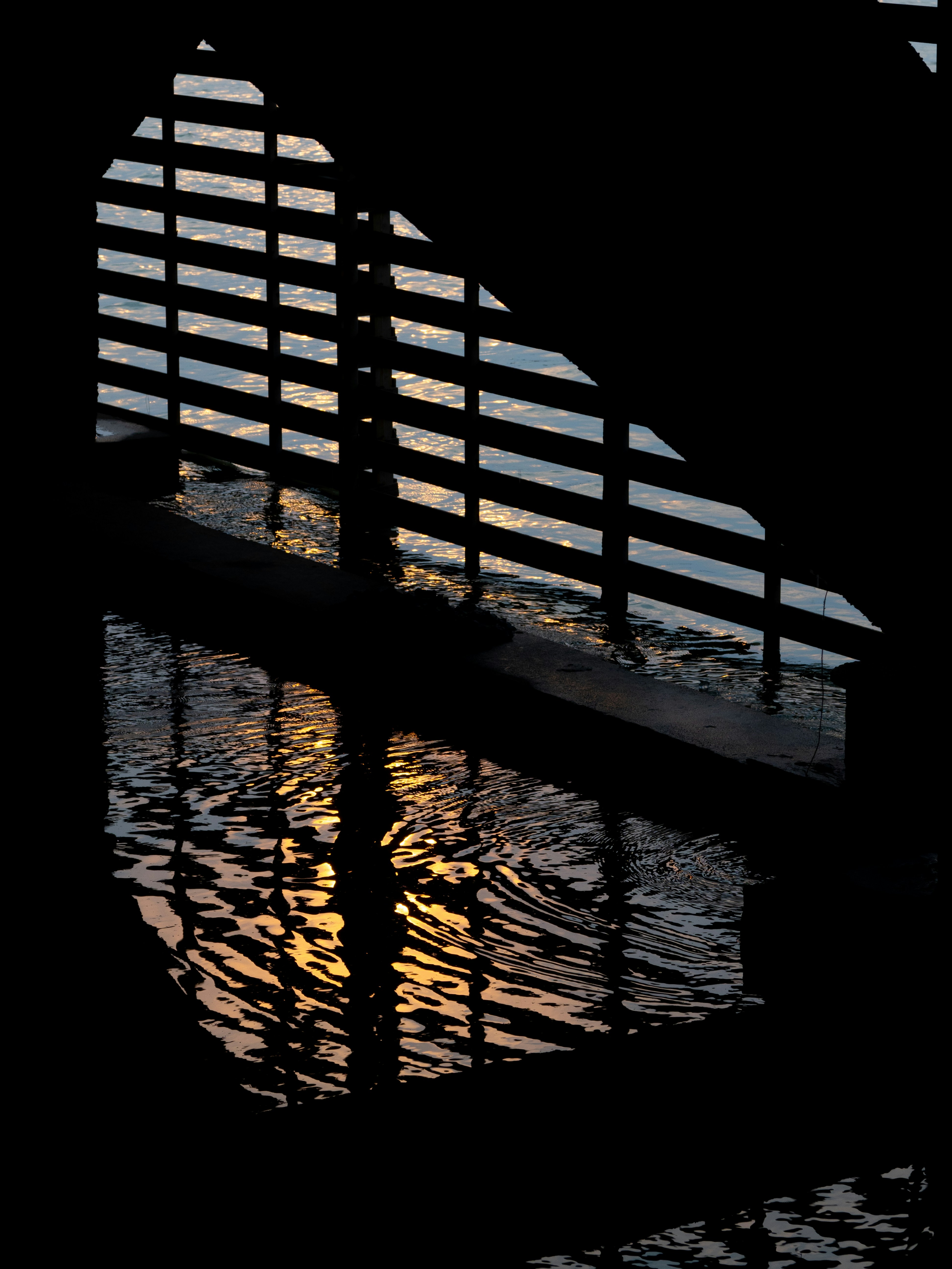 Silhouetted structure with intricate lattice casting shadows over rippling water, reflecting warm hues of sunset.
