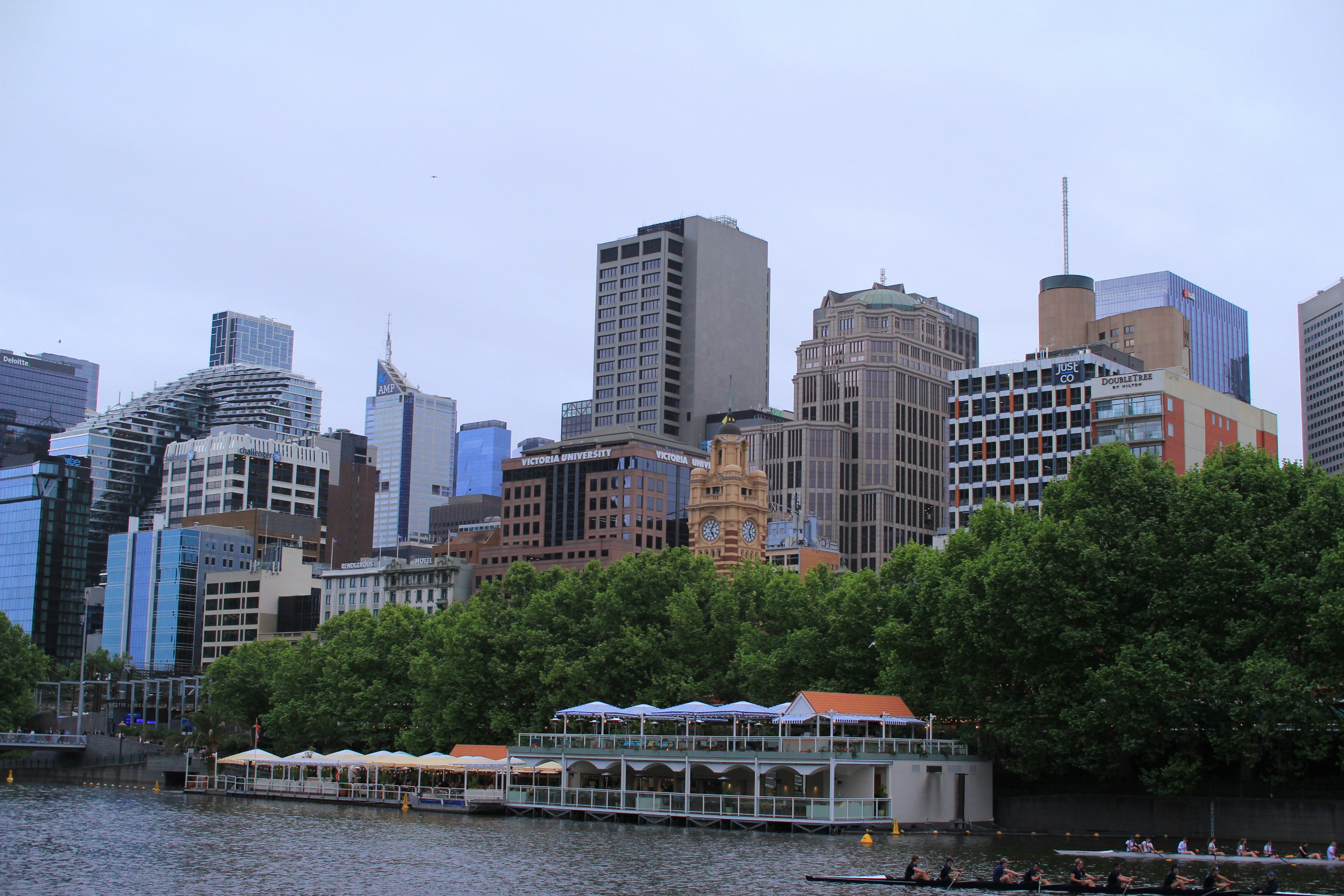 City skyline featuring a blend of contemporary buildings and lush greenery along the waterfront. A bustling atmosphere captures the essence of urban life.