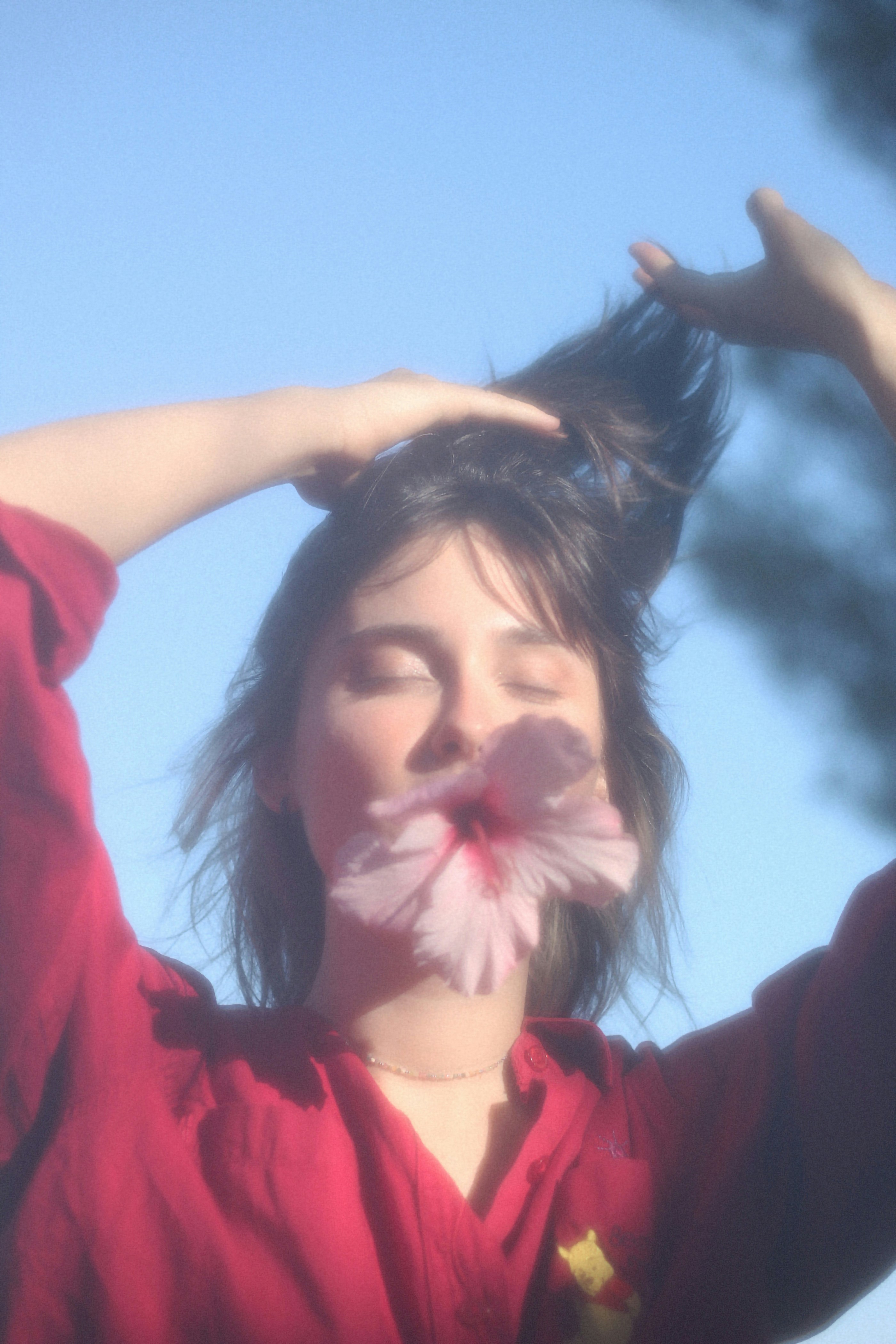 A young woman with closed eyes holds a hibiscus flower in her mouth, surrounded by soft blue skies. Her carefree pose and vibrant red shirt evoke a sense of freedom and connection with nature.