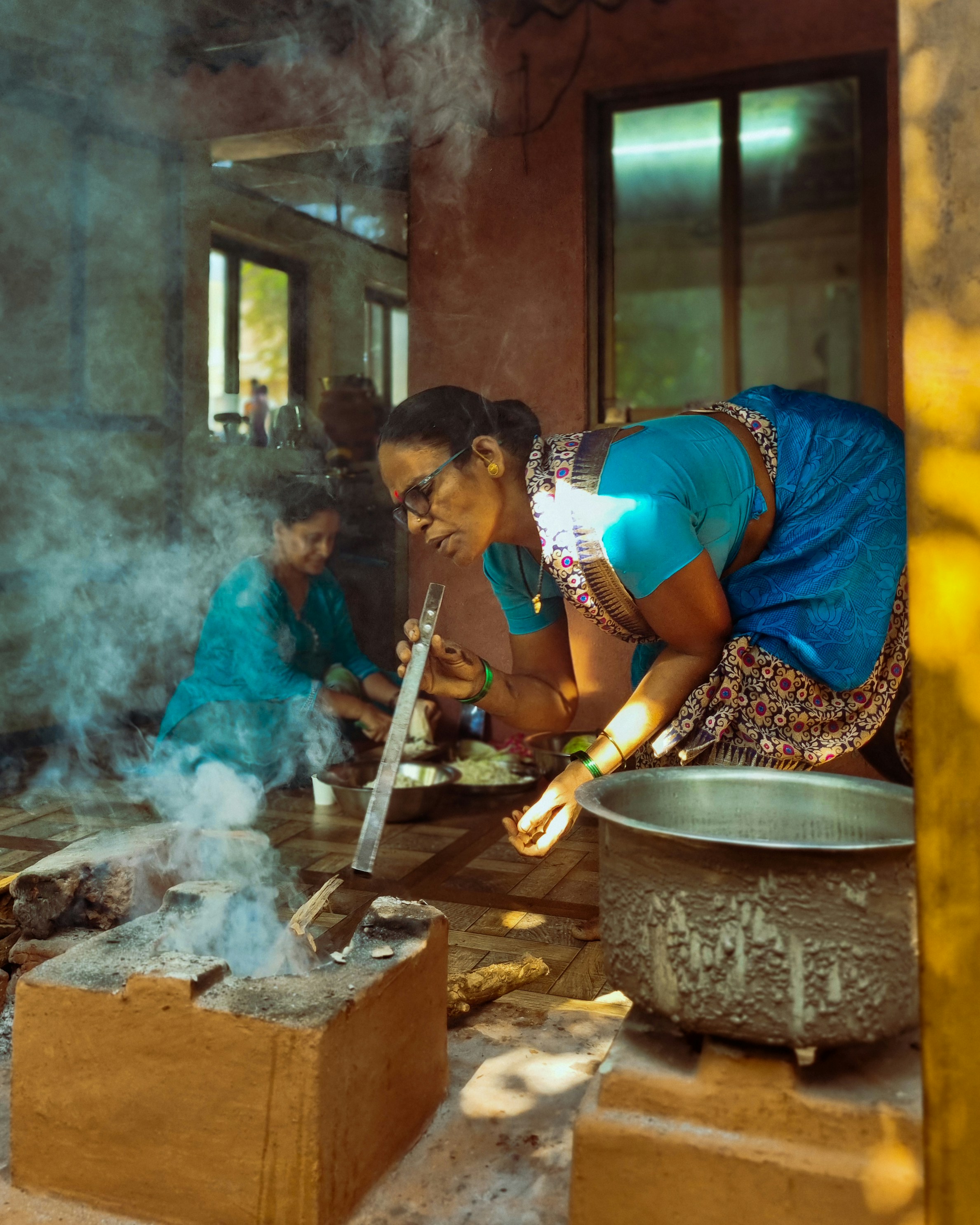 Woman in a vibrant blue saree tending to a traditional cooking fire, while another woman prepares food in the background. Smoke swirls around, adding depth to the scene.