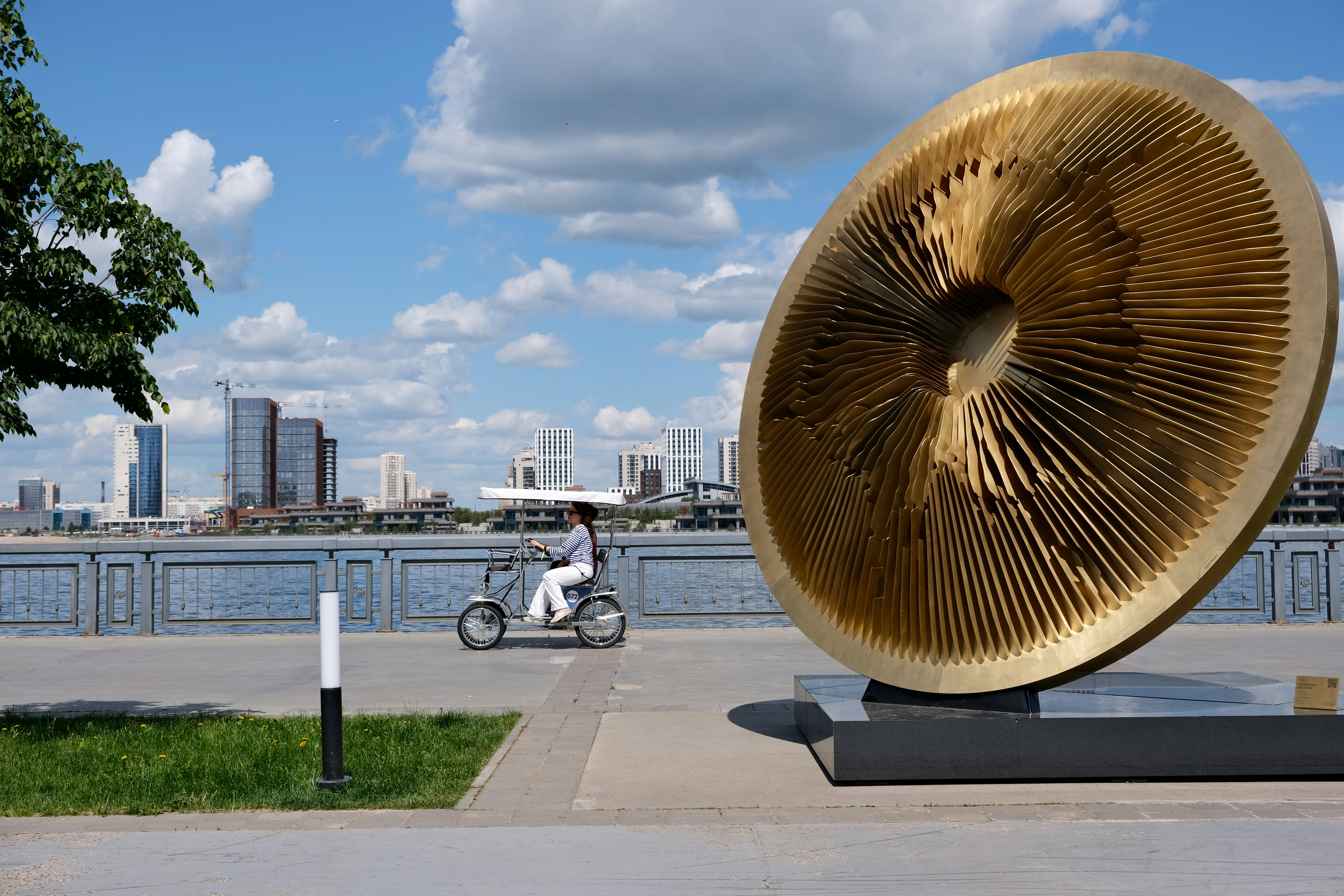A woman rides a tricycle near a large, golden, spiral-shaped sculpture by the waterfront, with a modern city skyline in the background. | A person rides by a bronze sculpture.