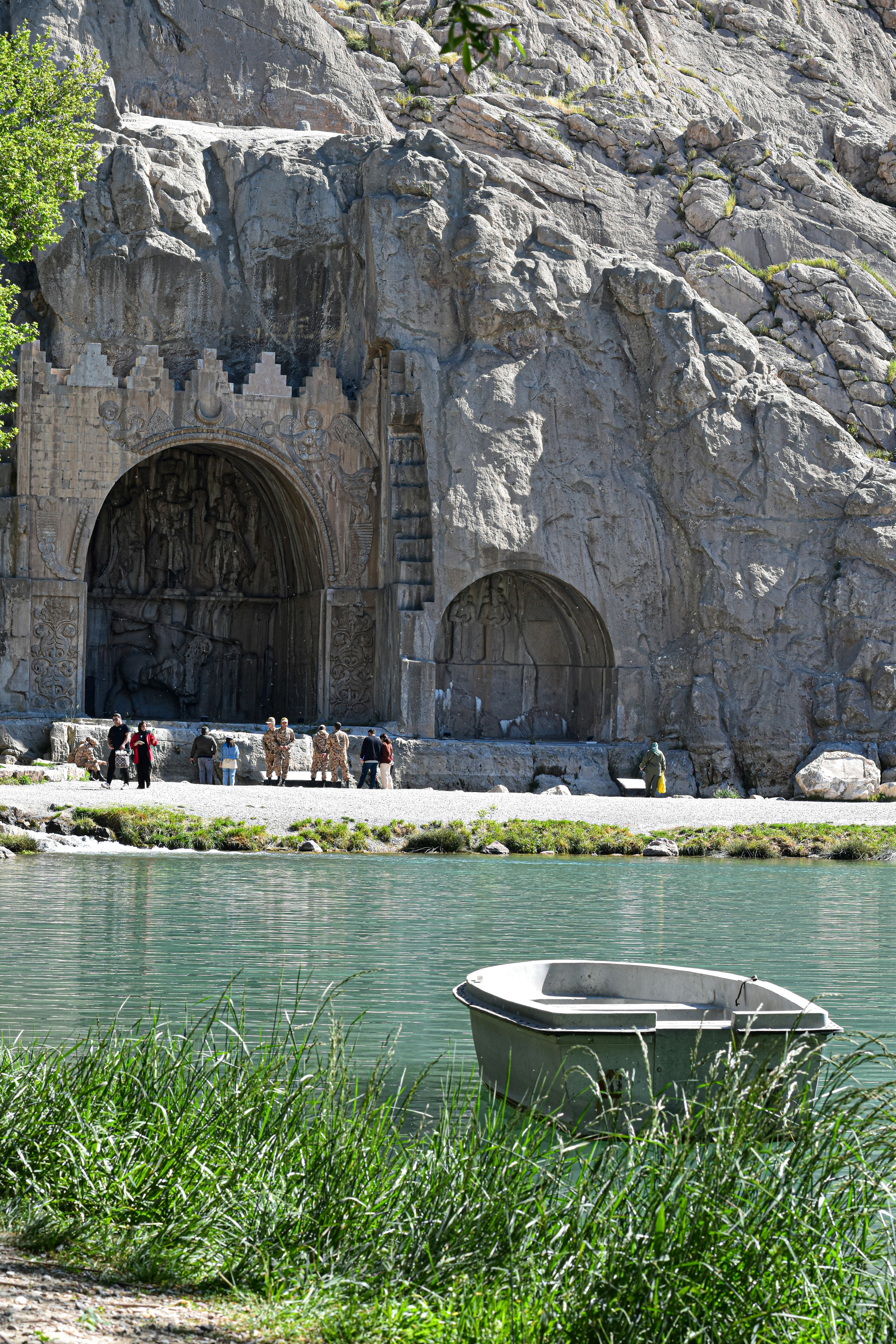 Intricate rock carvings nestled against a serene body of water, with visitors exploring the historical site nearby.