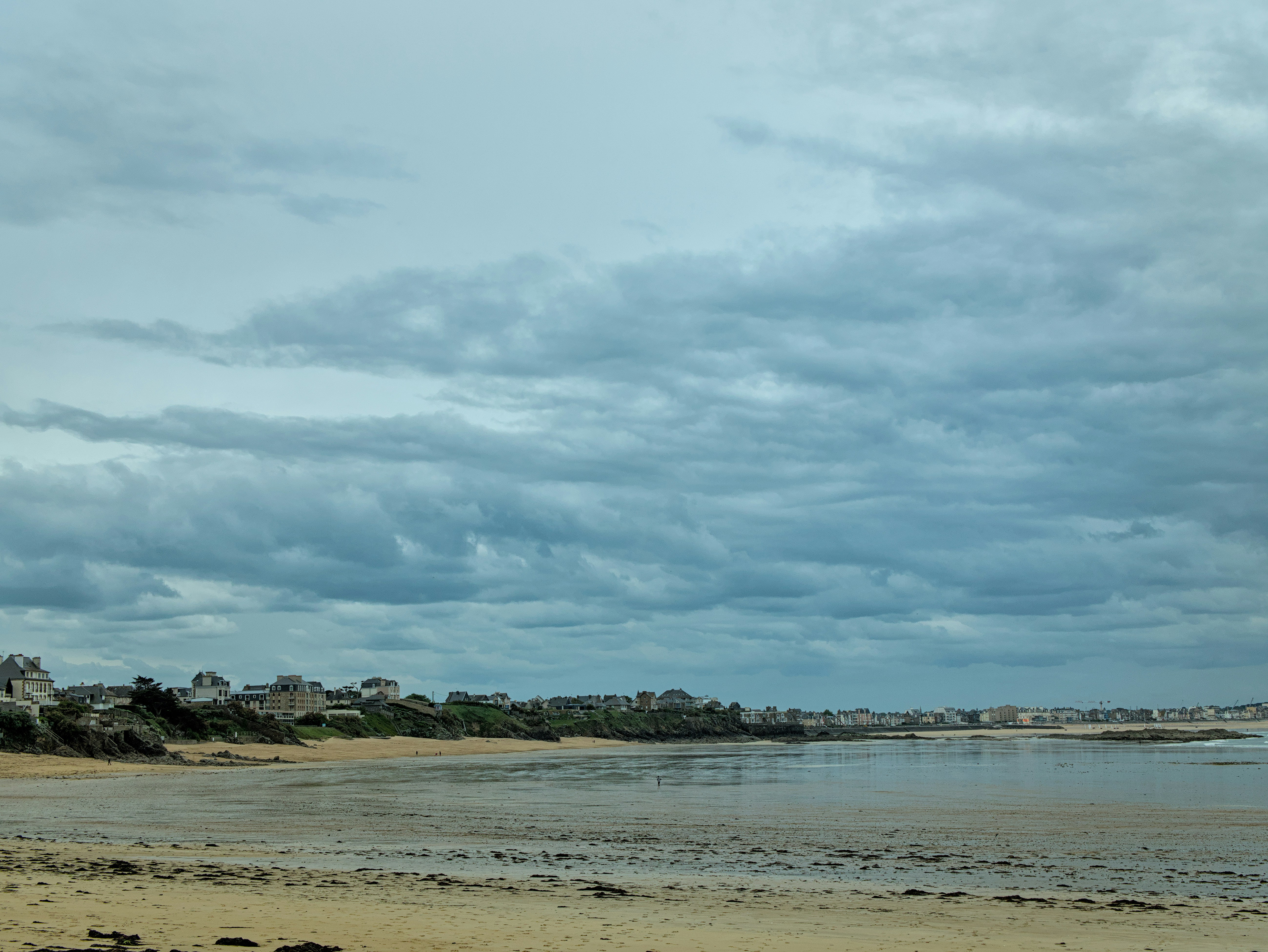 Serene beach landscape under a cloudy sky, showcasing the gentle curve of the shoreline and distant coastal homes.