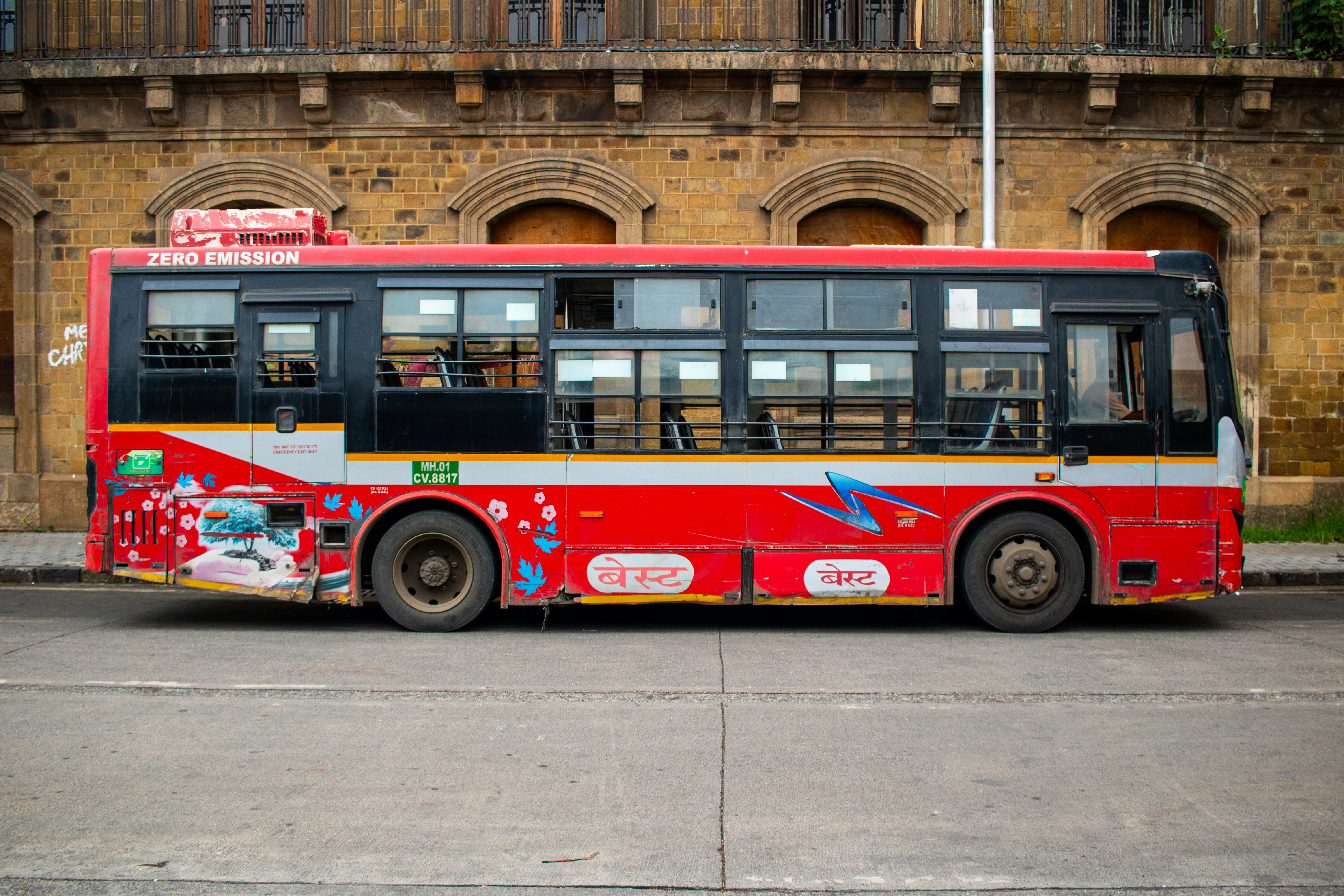 A red BEST bus passes through the neoclassical lanes of Ballard Estate, Mumbai—an area known for its British-era architecture and orderly street planning. The iconic bus, operated by the Brihanmumbai Electric Supply and Transport (BEST) undertaking, is a staple of daily commuting in the city. Its vibrant color contrasts with the muted stone façades of colonial buildings, creating a dynamic image of old meets new. This scene captures the essence of Mumbai: historic, functional, and always in motion.