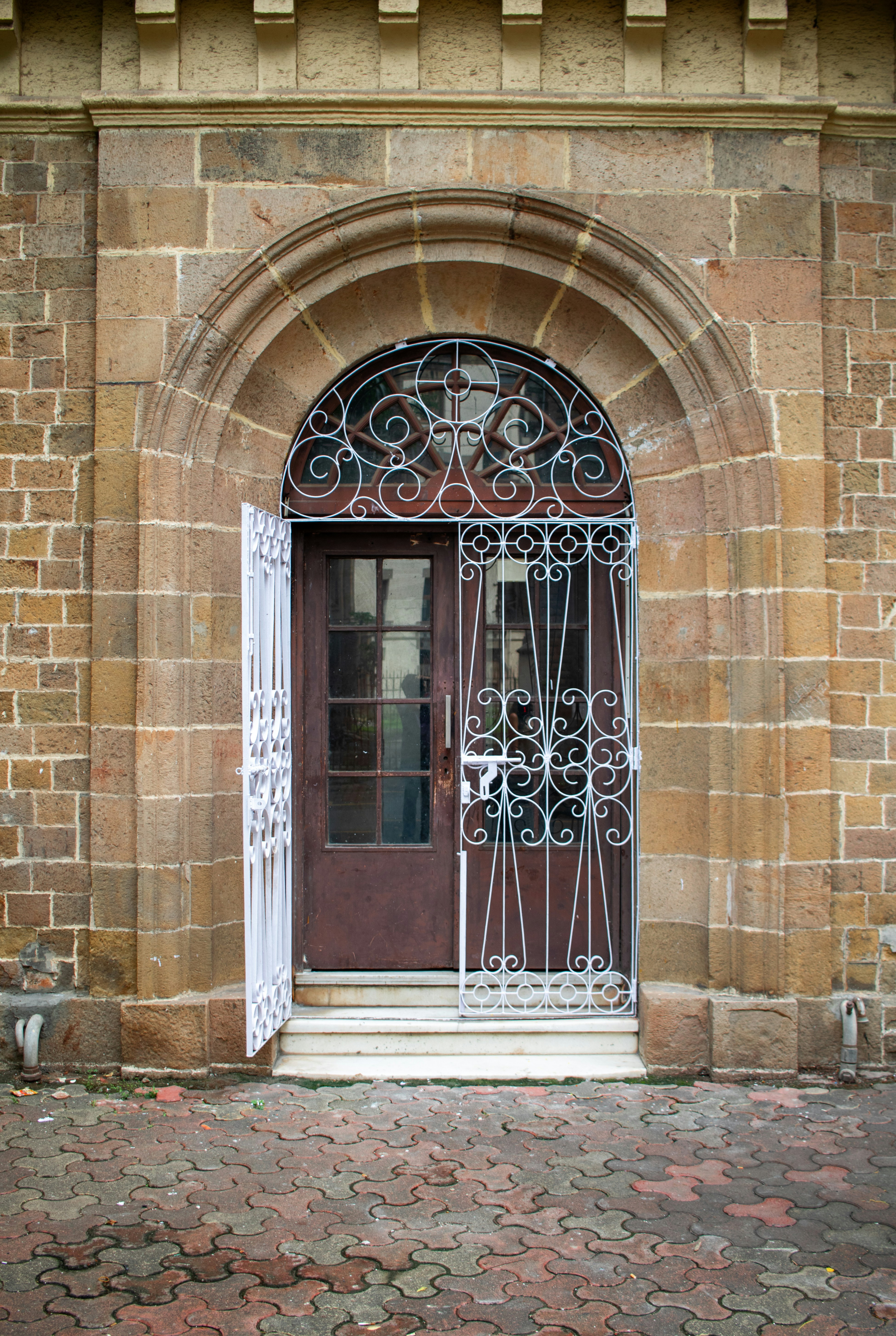 An elegant arched doorway in Ballard Estate, Mumbai, showcases the area’s colonial architectural legacy. Framed in carefully laid brownstone, the entrance features intricate white wrought ironwork guarding a weathered wooden door. This ornate design is characteristic of the British-era heritage buildings that populate South Mumbai’s historic business district. | A brown door has an ornate gate in front.