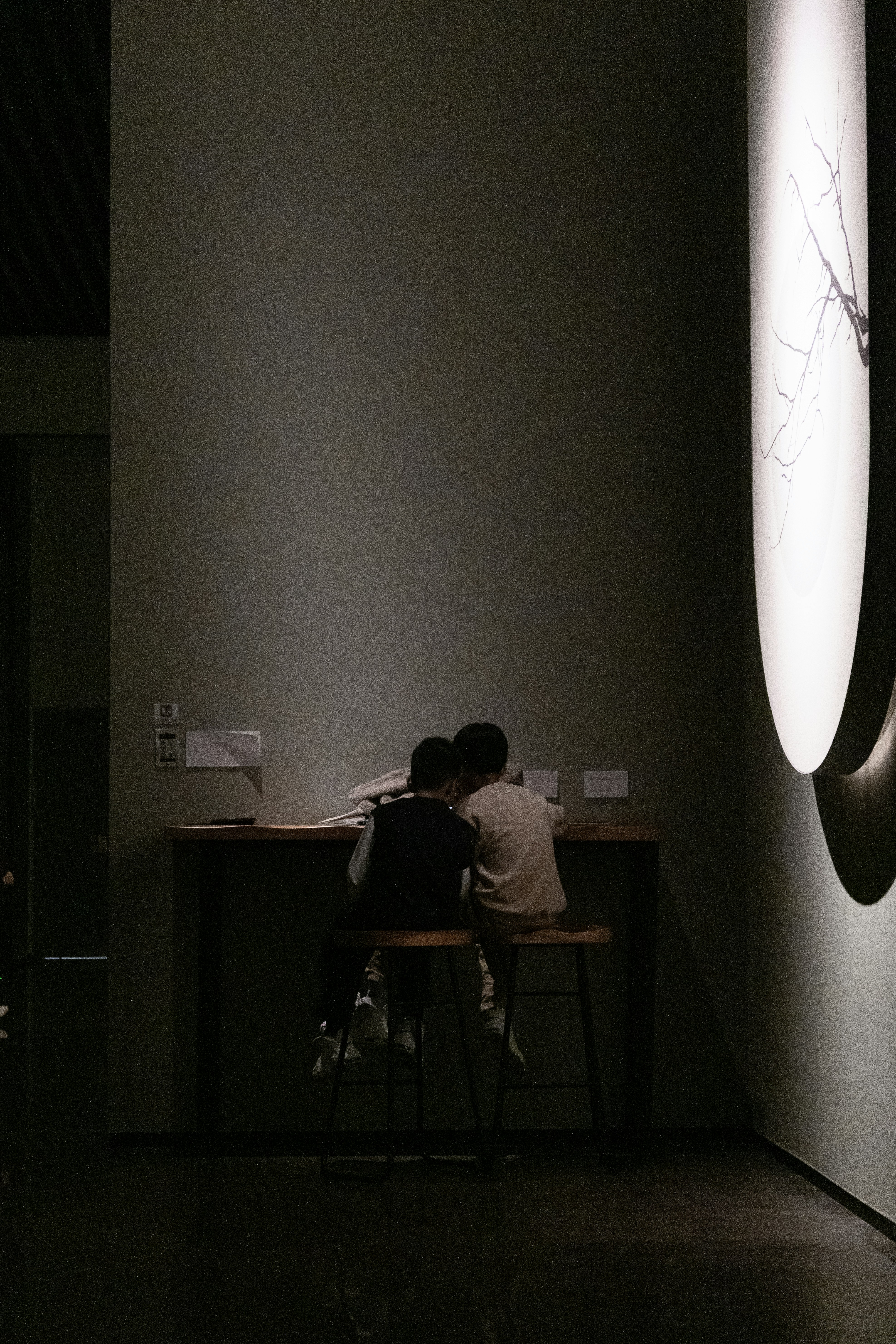 Two children share a quiet moment at a sleek bar table, illuminated by soft light against a minimalistic wall adorned with a striking shadow.