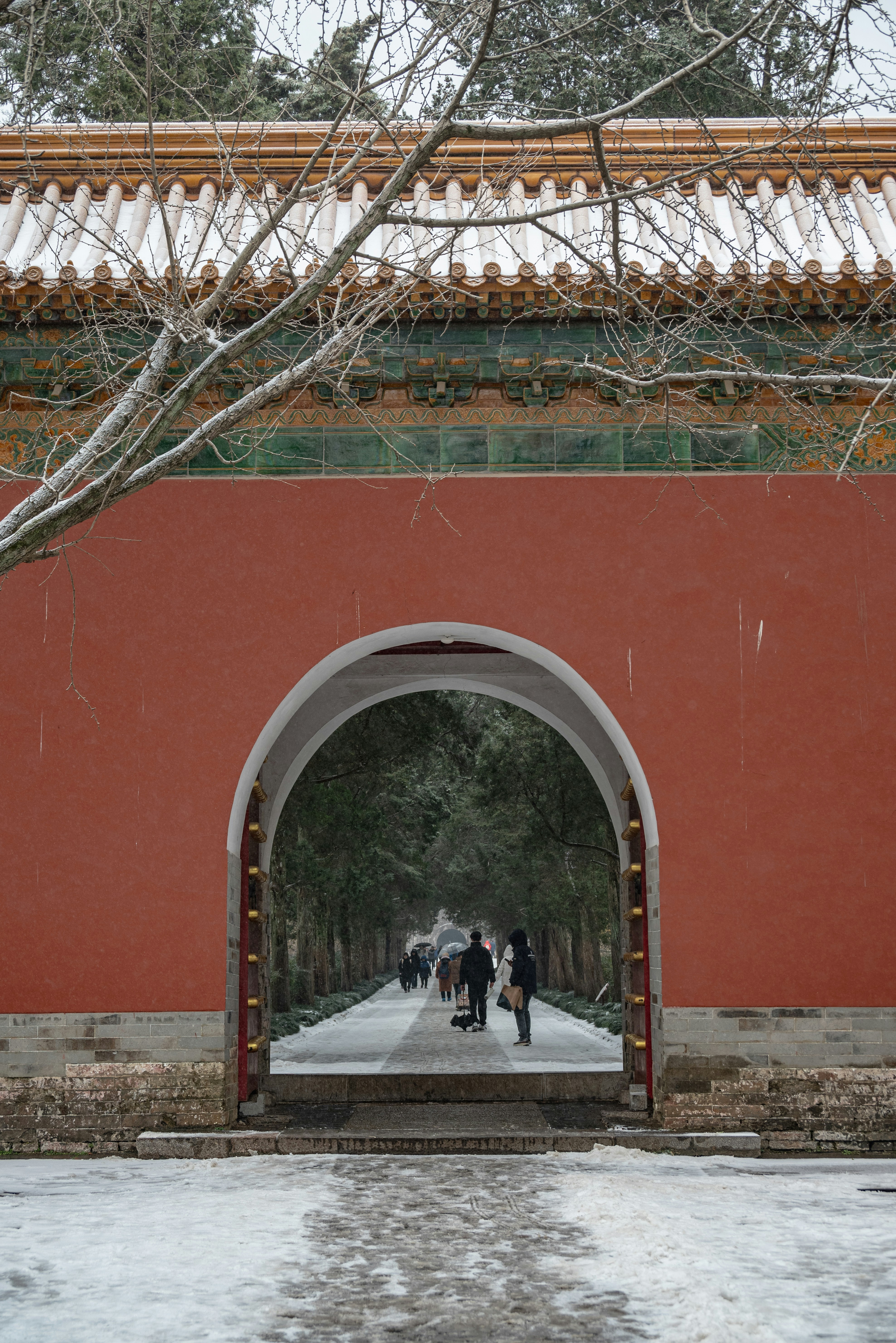 Archway leading to a snow-covered path, framed by trees and a vibrant red wall. Visitors stroll through the serene landscape.