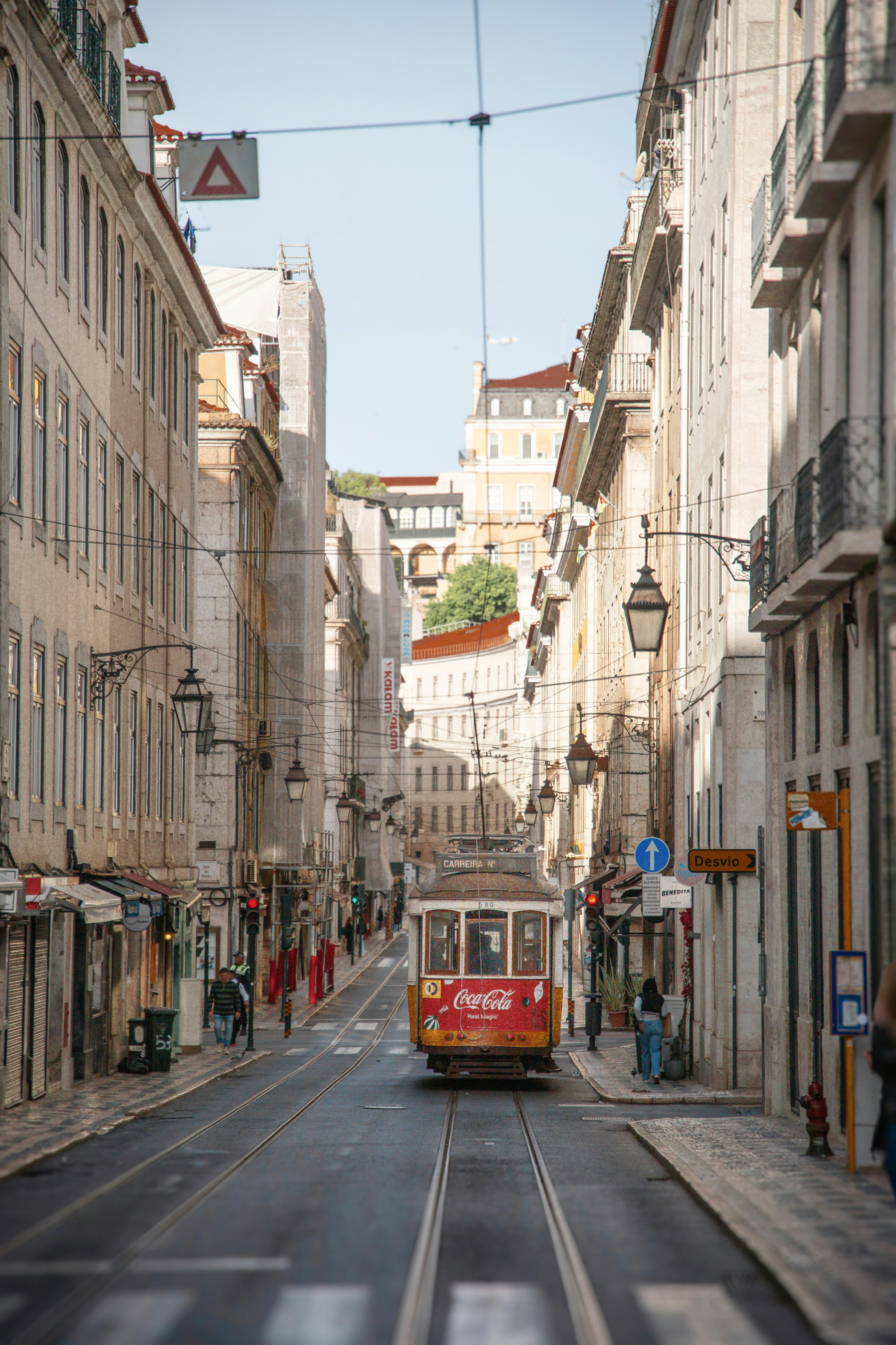 A vibrant yellow tram navigates through a narrow Lisbon street lined with traditional architecture and quaint shops. The scene captures the essence of urban life in Portugal.