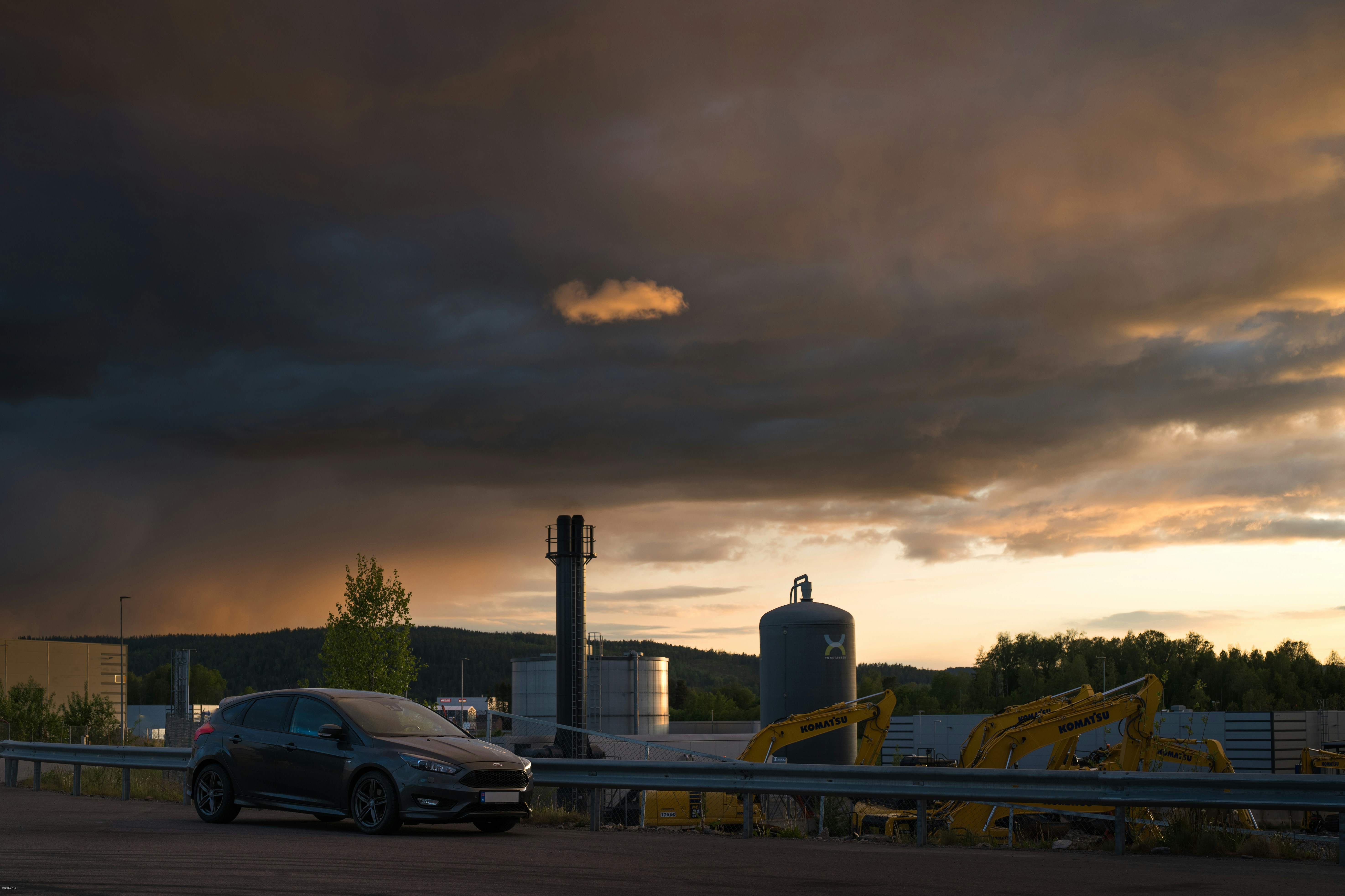 A sleek gray car parked beside industrial machinery under a dramatic sky filled with dark clouds and a hint of sunset glow.