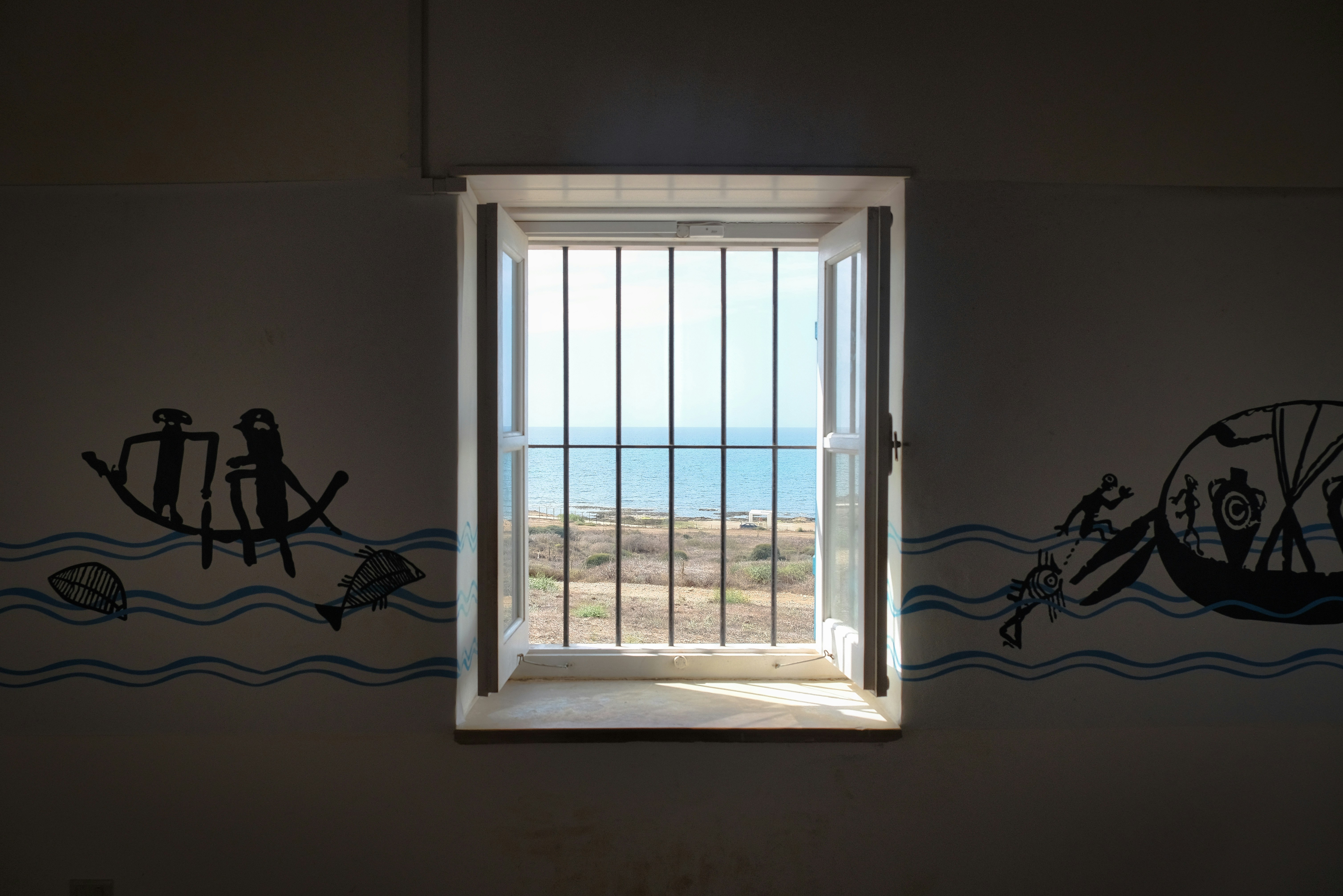 View of the Mediterranean Sea through a barred window in a seaside building, with playful maritime-themed murals decorating the interior walls.