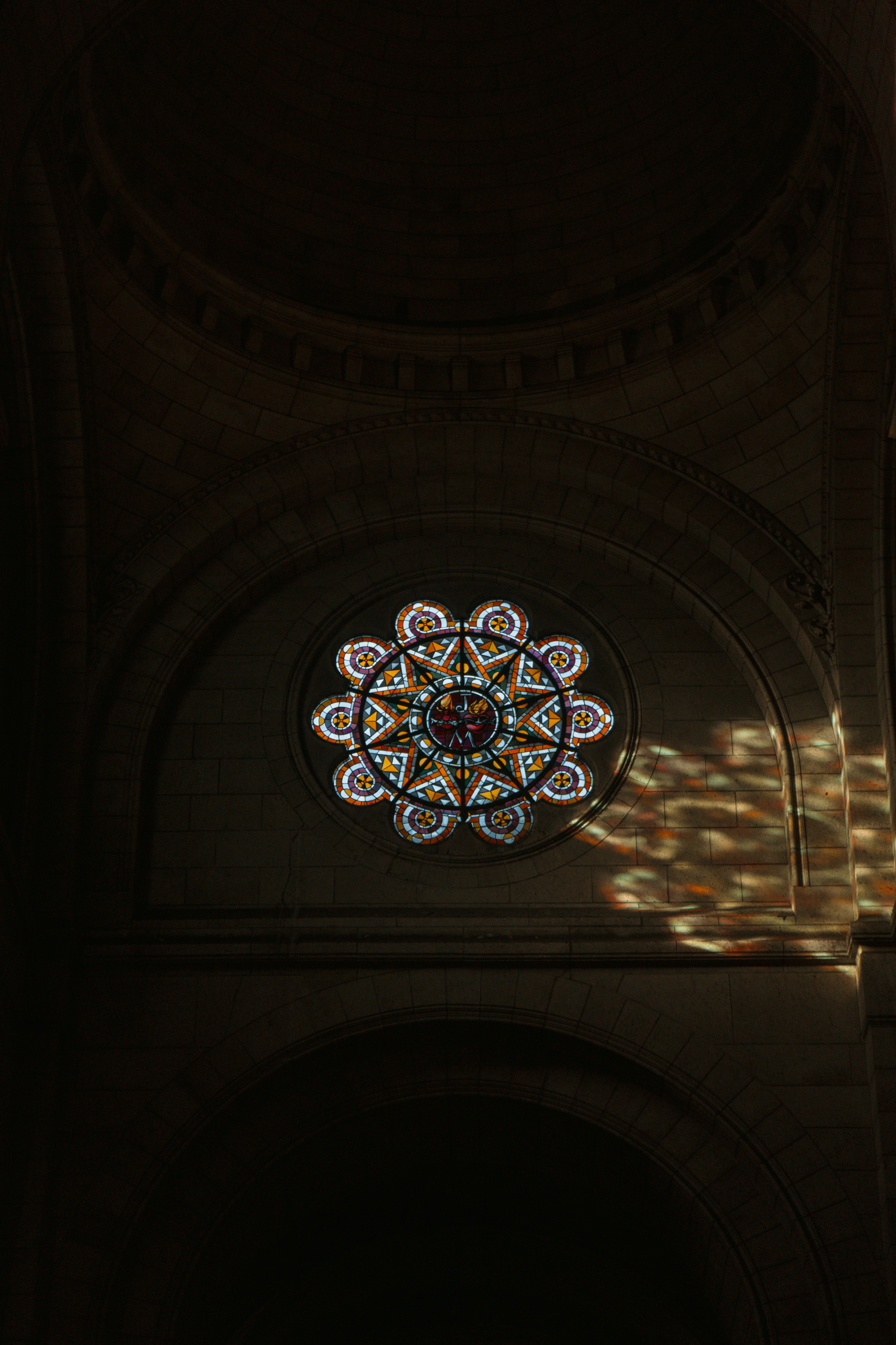 Intricate stained glass window casting colorful reflections on the stone wall of a dimly lit space.