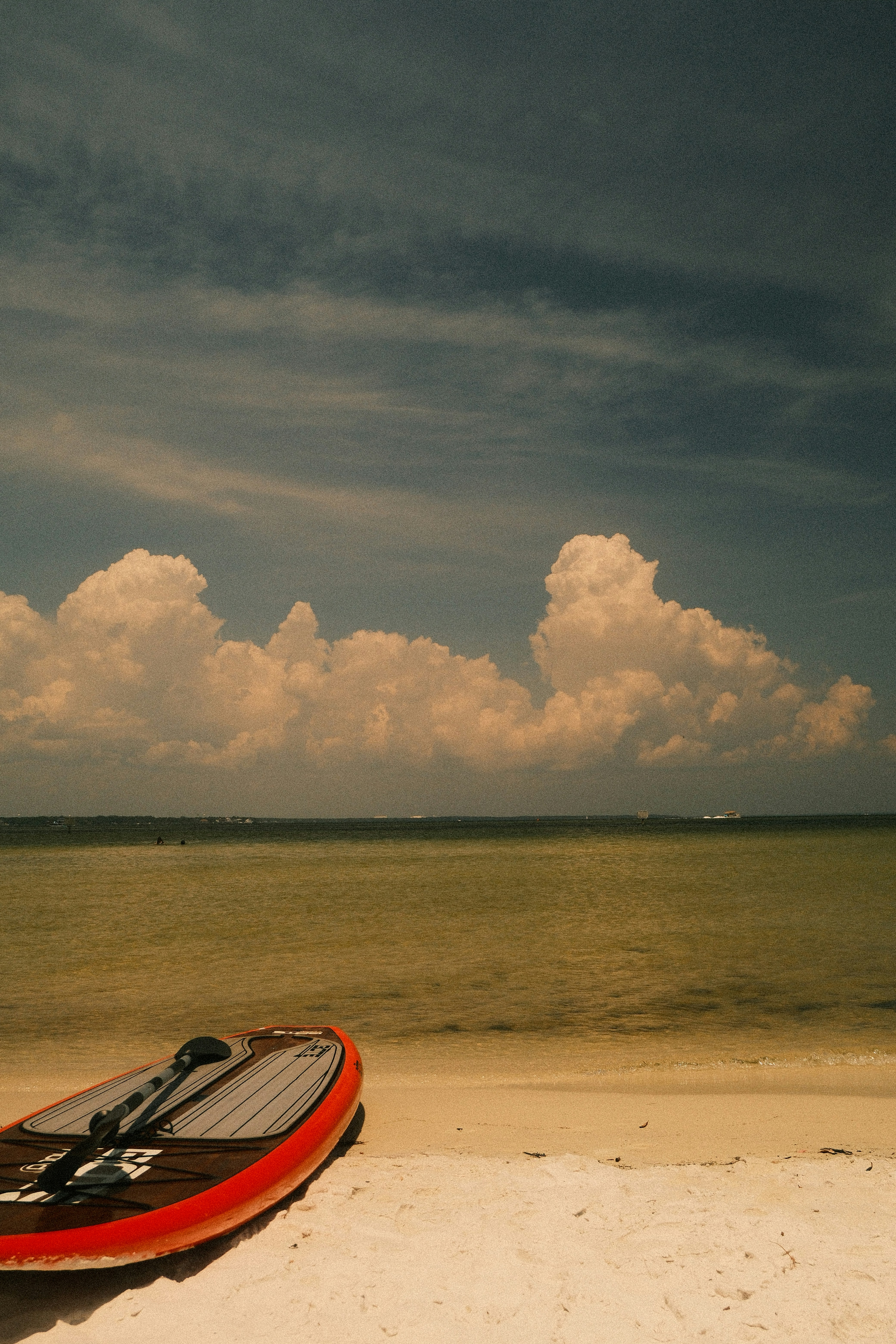 A vibrant paddleboard rests on the sandy beach as fluffy clouds drift above a tranquil sea. The scene evokes a sense of calm and leisure.