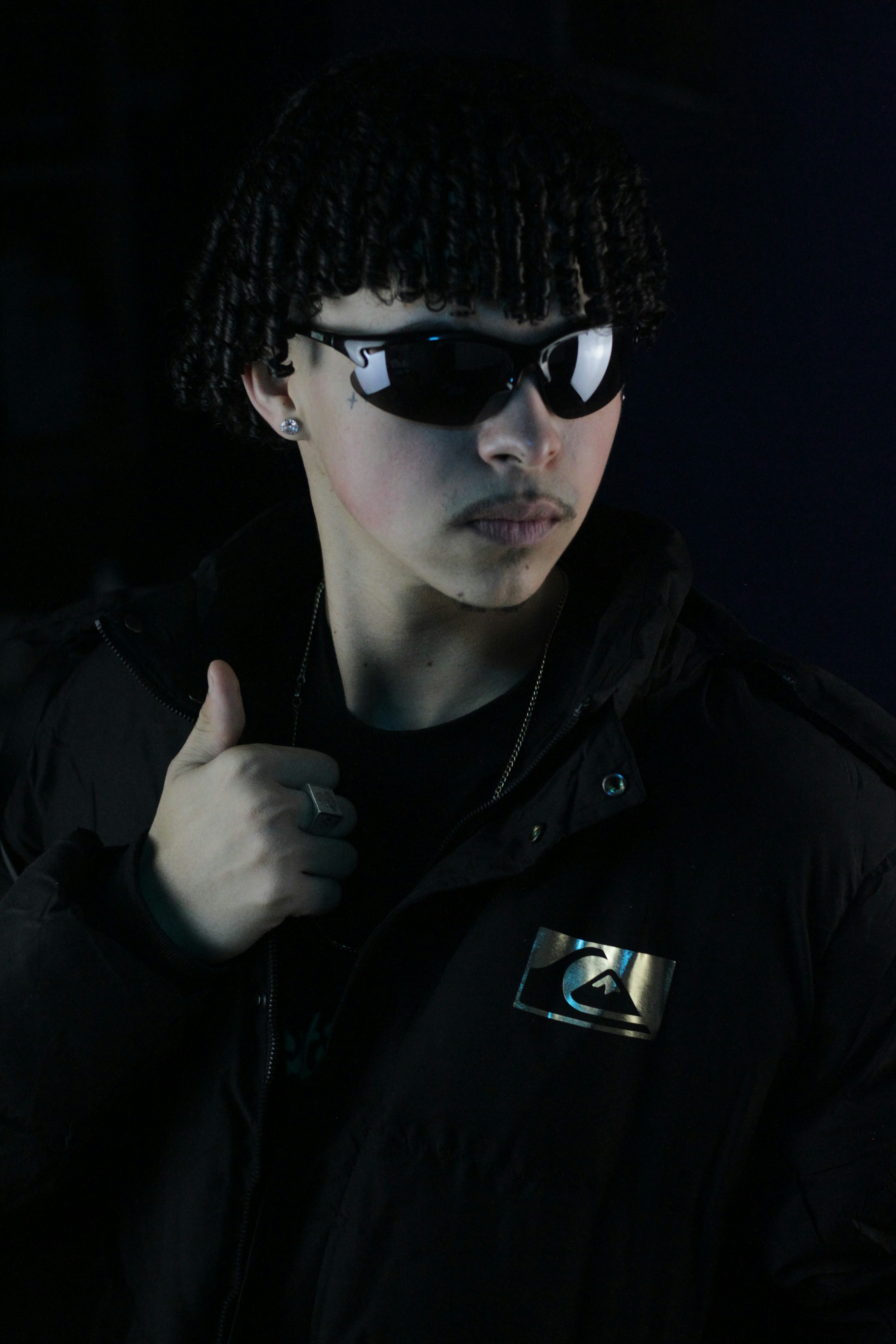 A young man in a stylish black puffer jacket and sunglasses strikes a confident pose, exuding urban flair against a dark background.