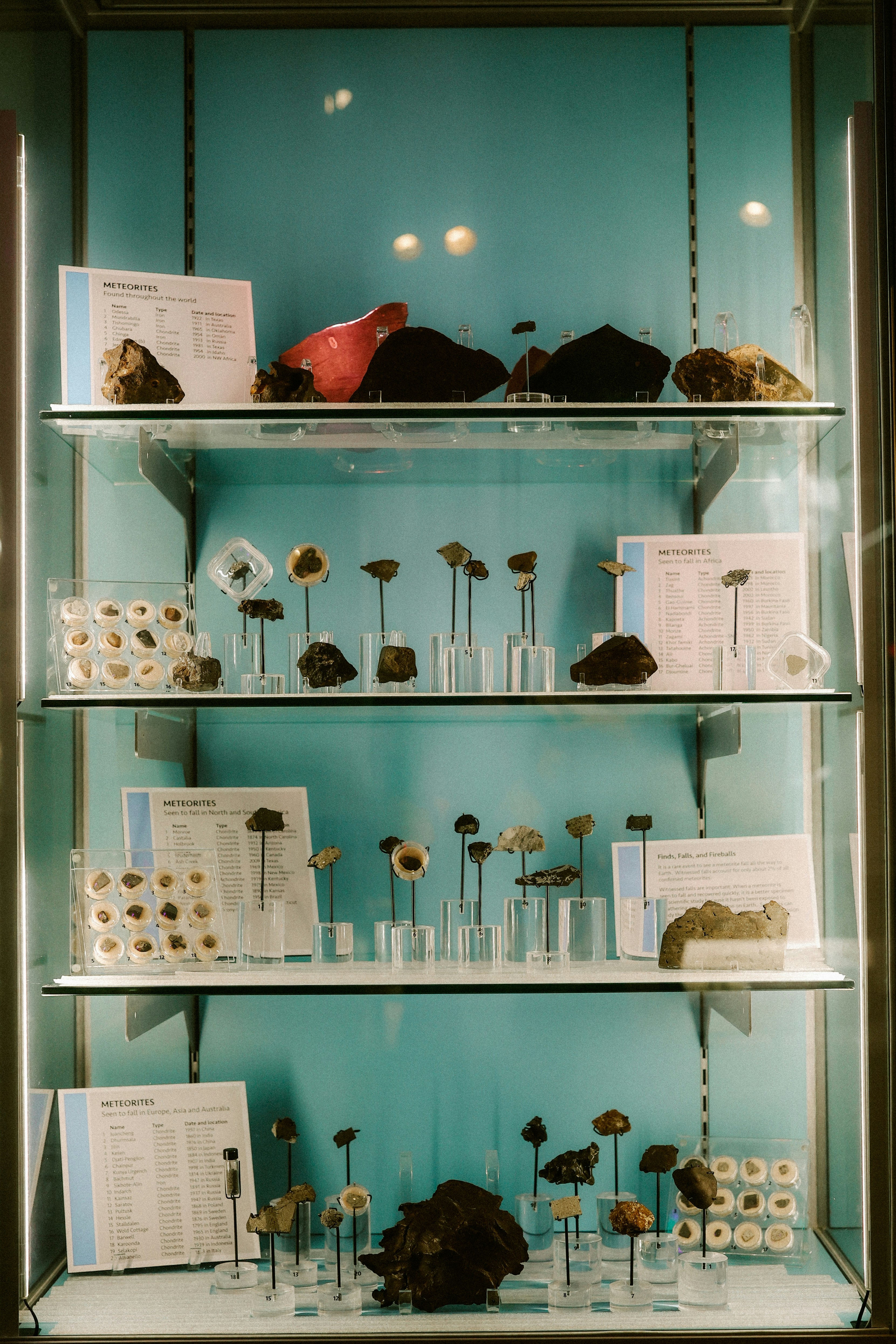 Display of various meteorites in a glass case, featuring informative cards and unique specimens arranged on clear stands.