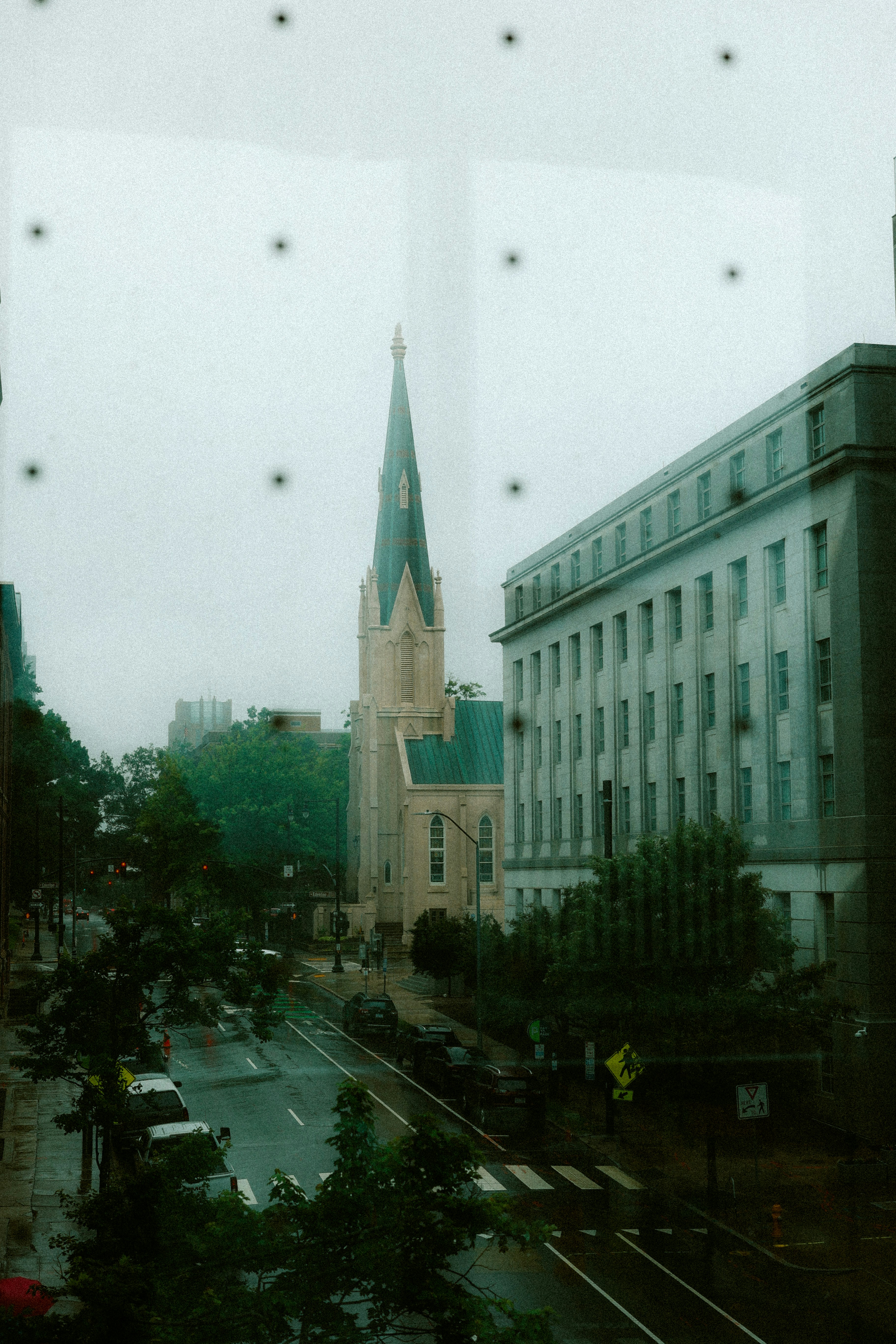 Historic church steeple rises amidst a misty urban landscape, framed by modern buildings and wet streets.