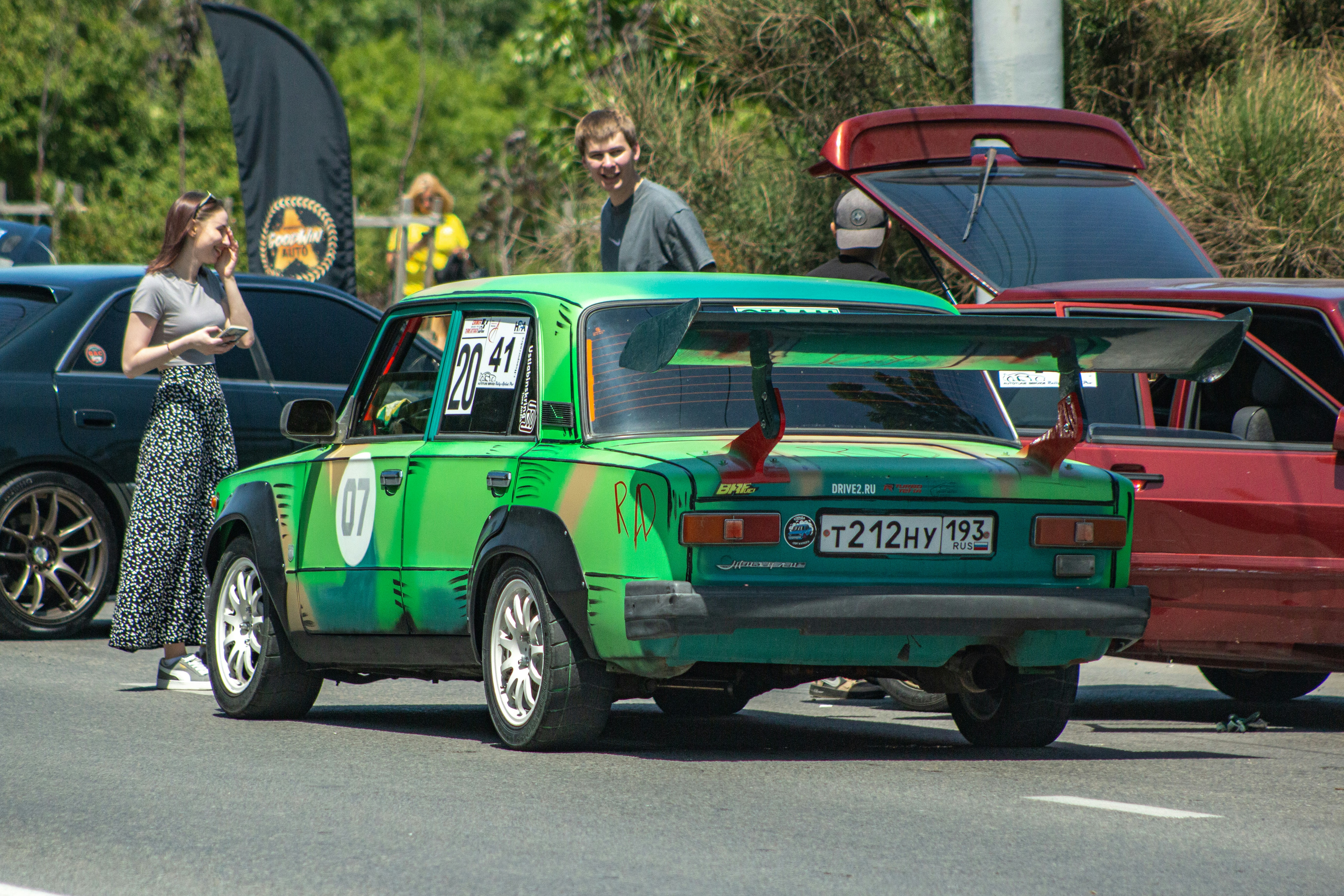 Green race car with open rear hatch at a daytime car meet