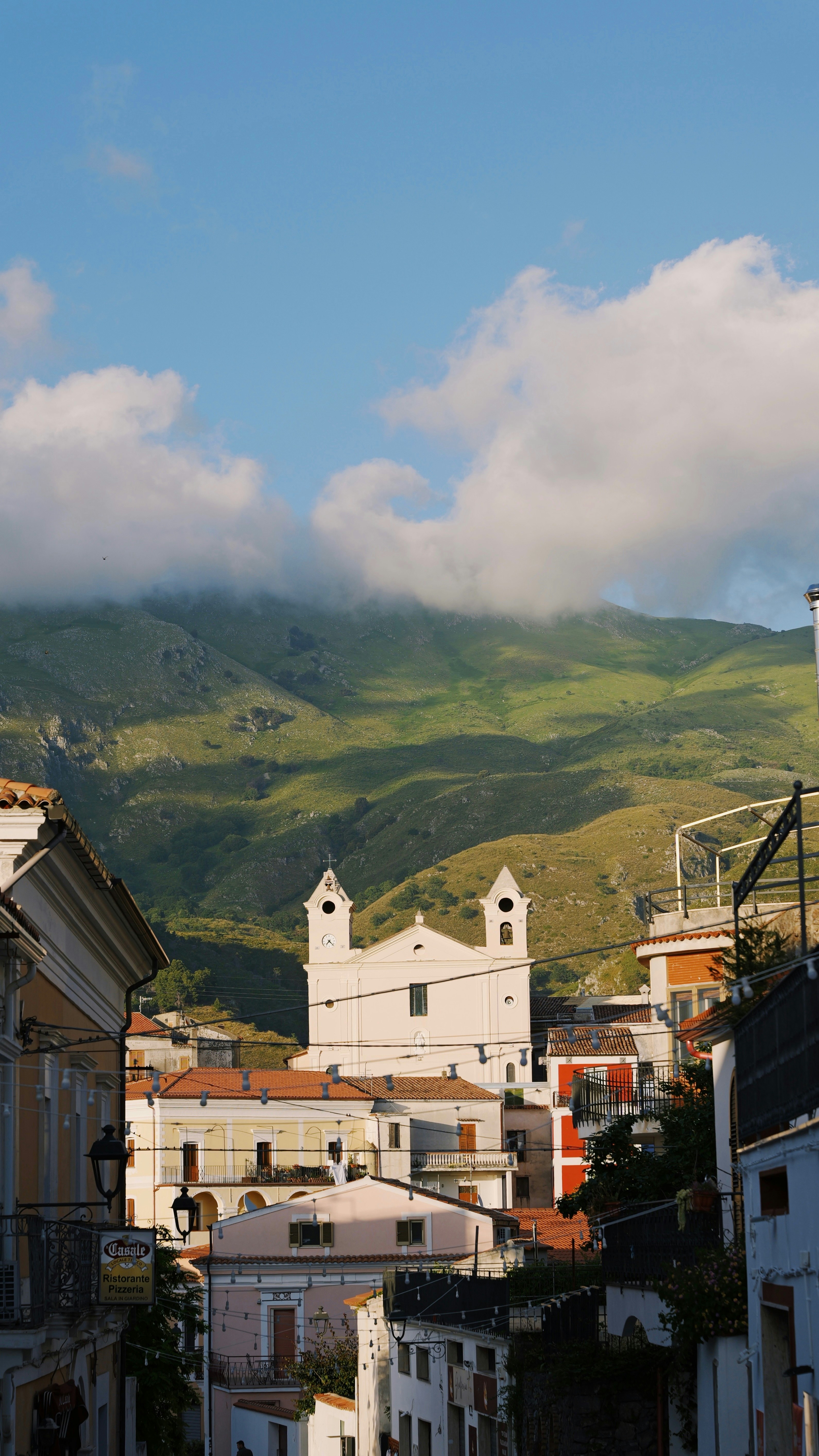 Charming village nestled in the foothills, showcasing traditional architecture against a backdrop of rolling green mountains. Soft clouds drift in the blue sky.