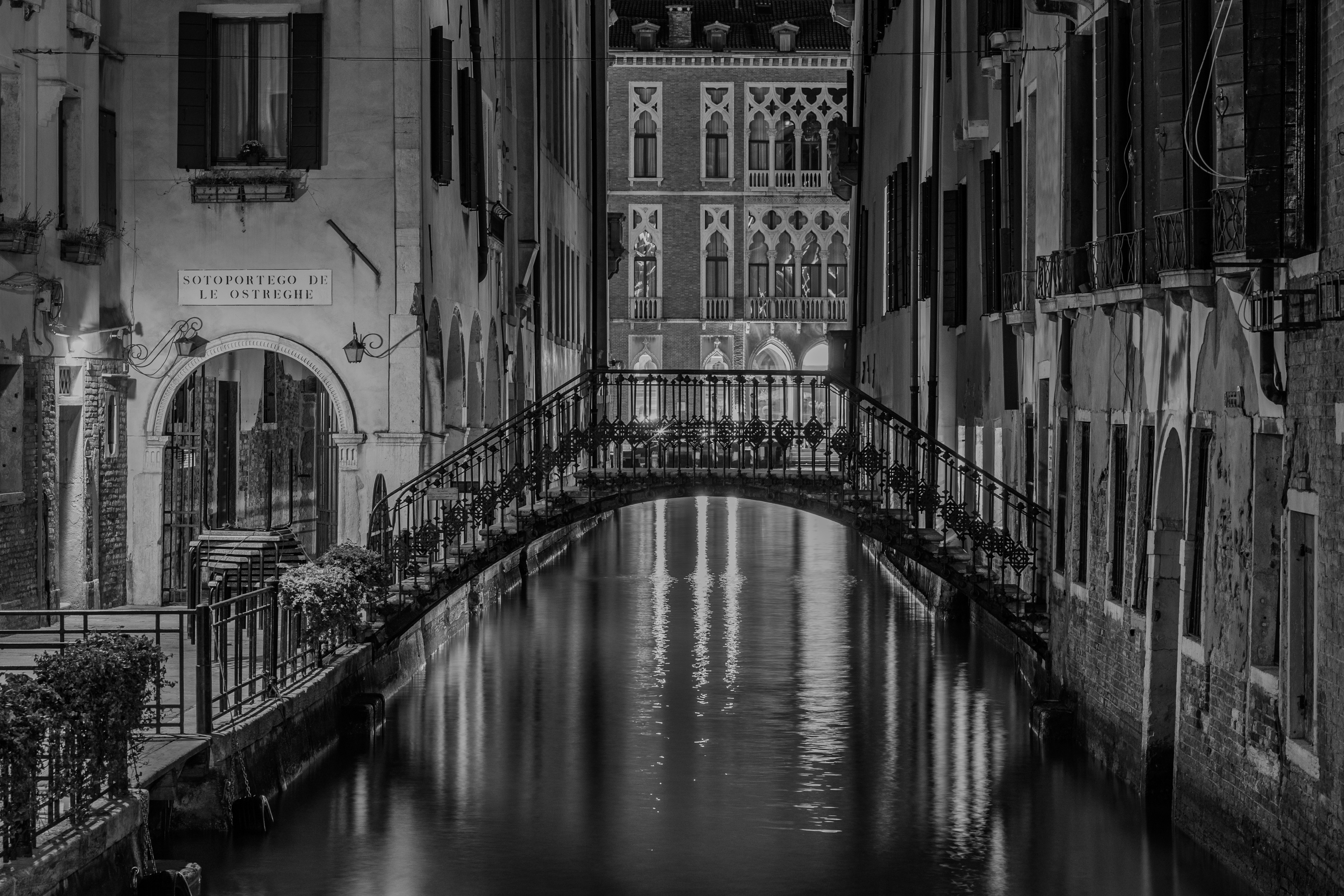 A serene canal scene in Venice featuring a quaint bridge and historic architecture reflected in the still water. The monochrome tones enhance the timeless quality of the setting.