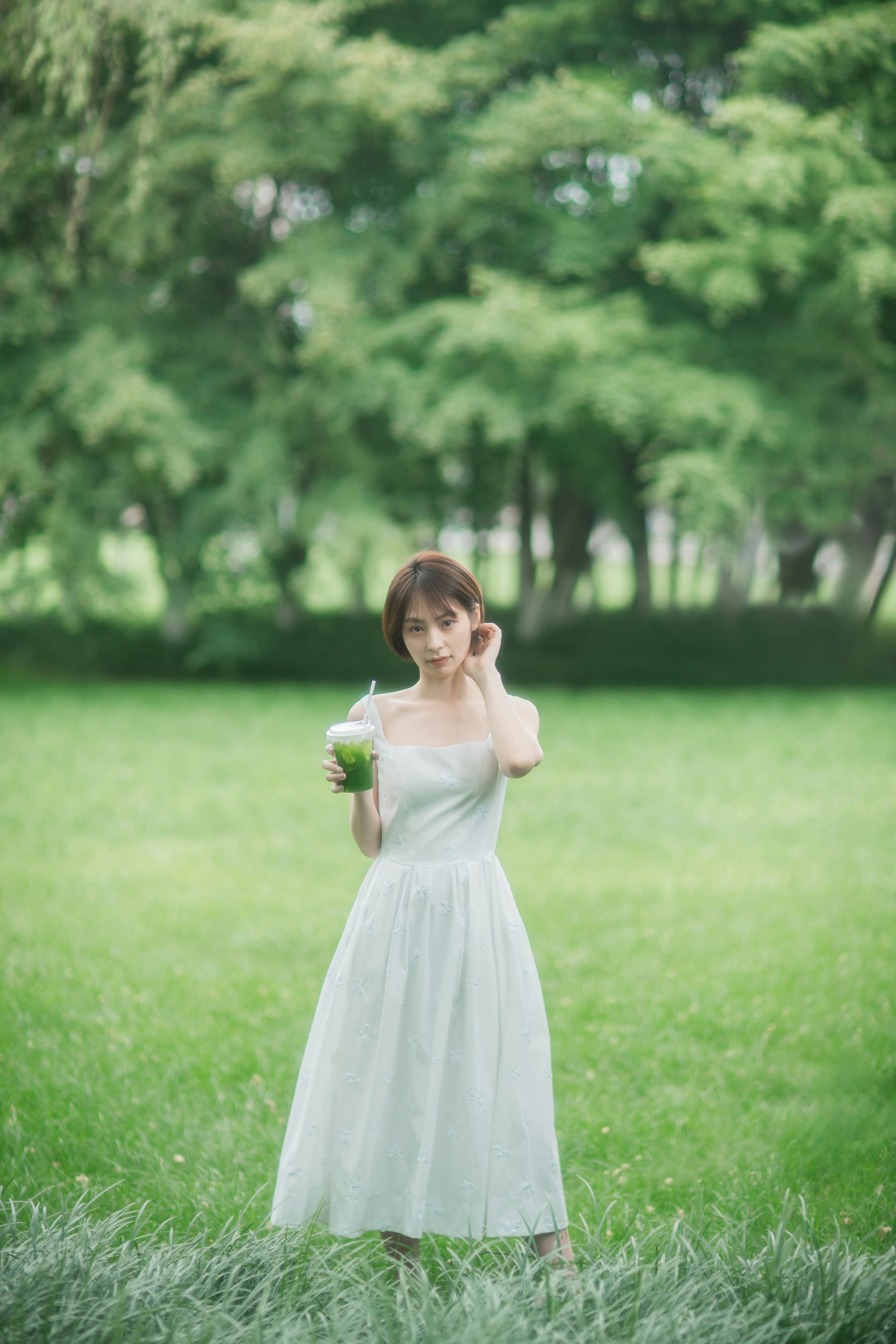 Young woman in a white dress holding a green beverage, standing in a lush green field surrounded by trees.