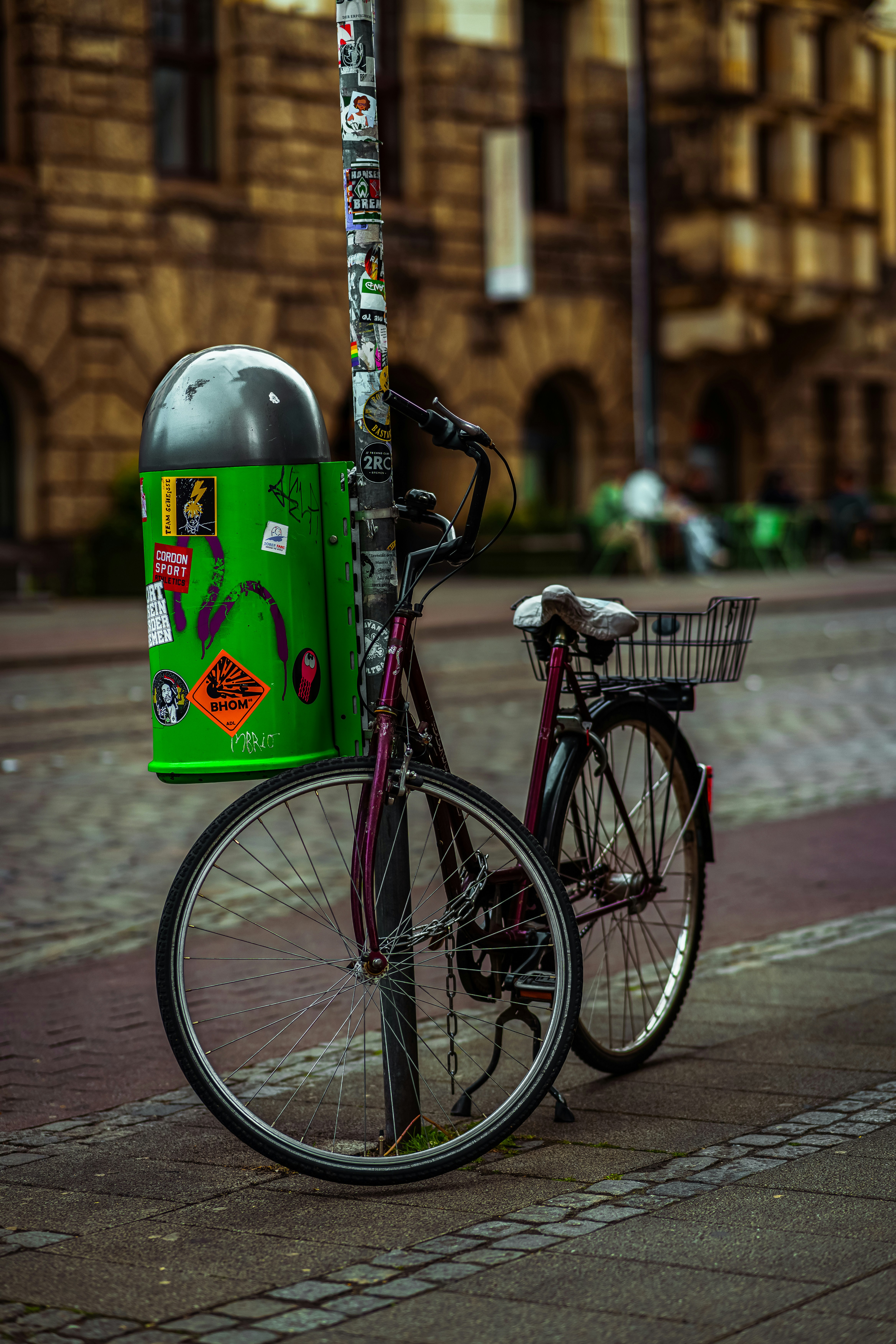 Maroon City Bicycle Leaning by a Stickered Post on a Cobblestone Street