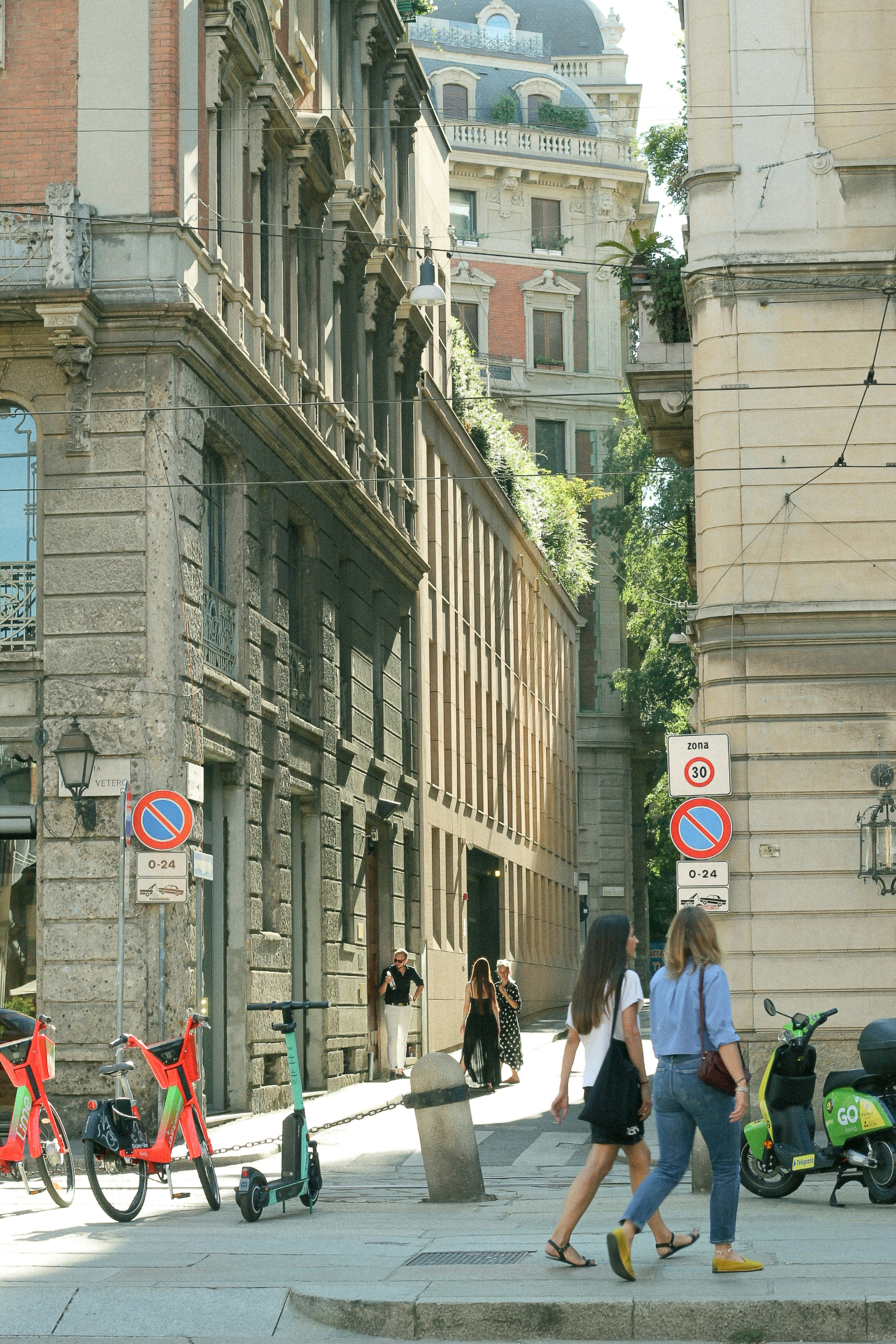 Two pedestrians stroll past modern e-scooters and historic architecture in a lively urban setting.