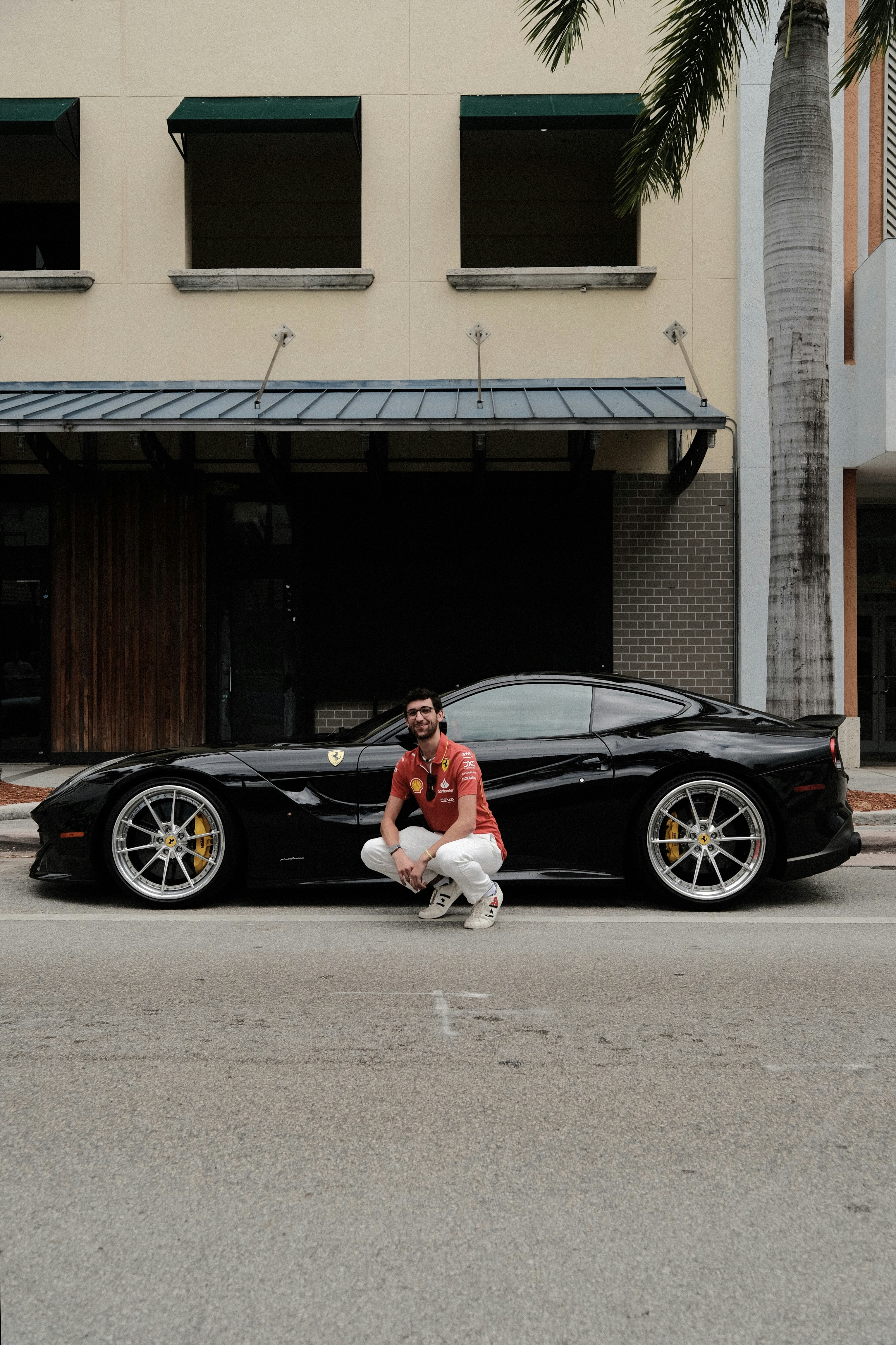 A man in a red shirt kneels beside a sleek black sports car, showcasing its striking design and intricate details. The urban backdrop enhances the scene's modern vibe.