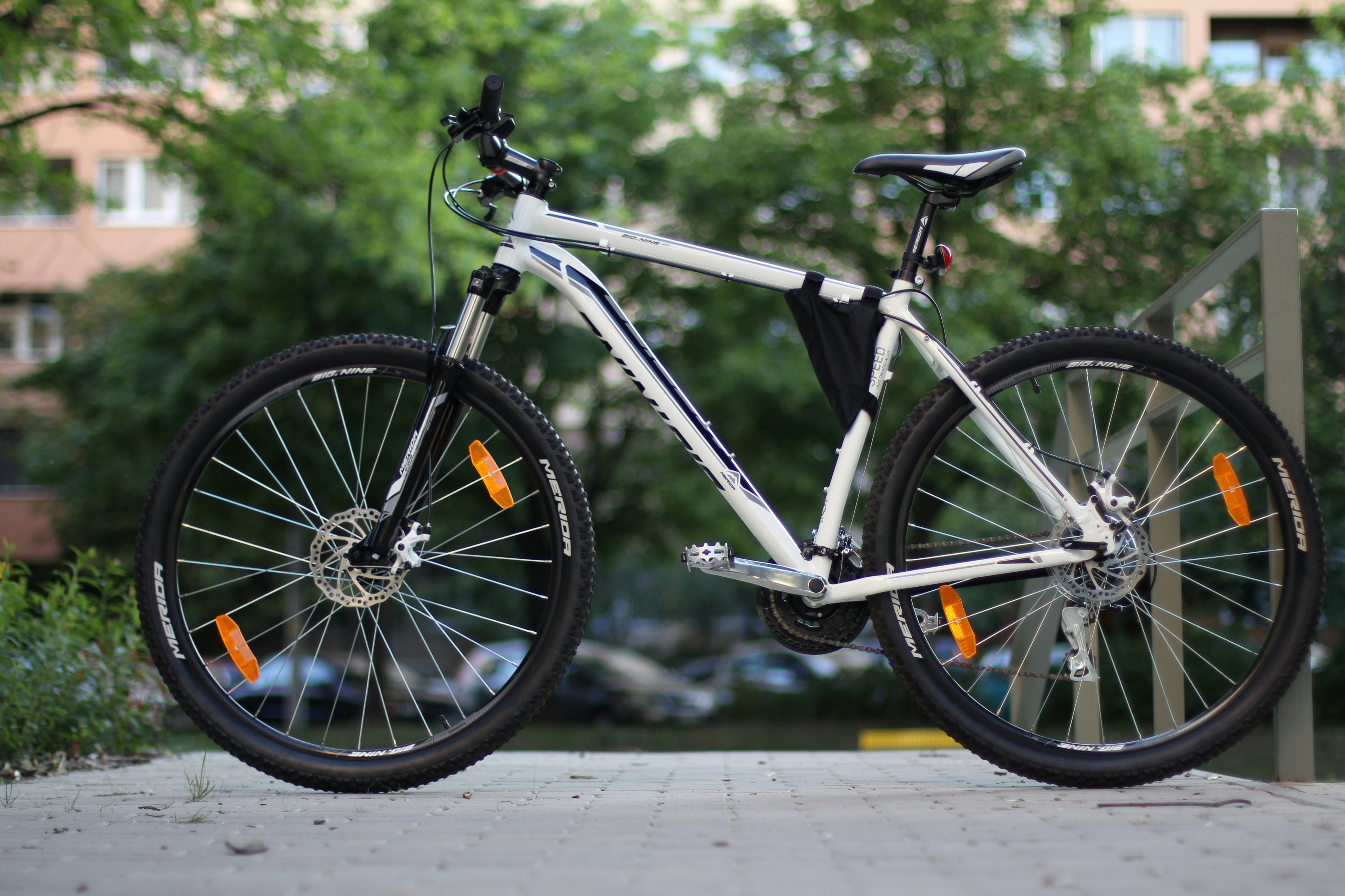 Mountain bike positioned on a paved path with a blurred background of greenery and buildings, highlighting its design and readiness for exploration.