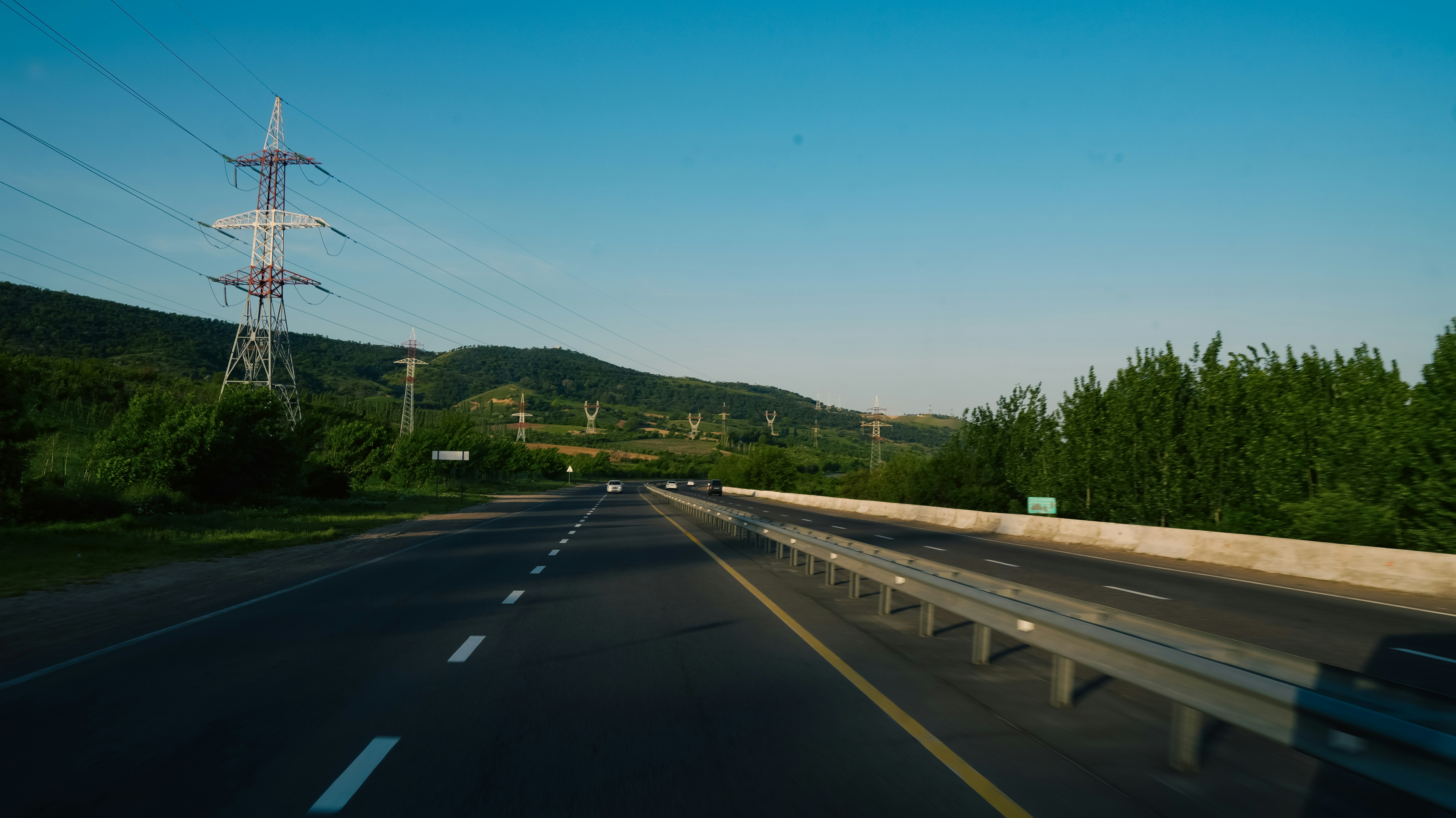 A highway stretches into the distance, flanked by lush greenery and towering power lines under a clear blue sky.