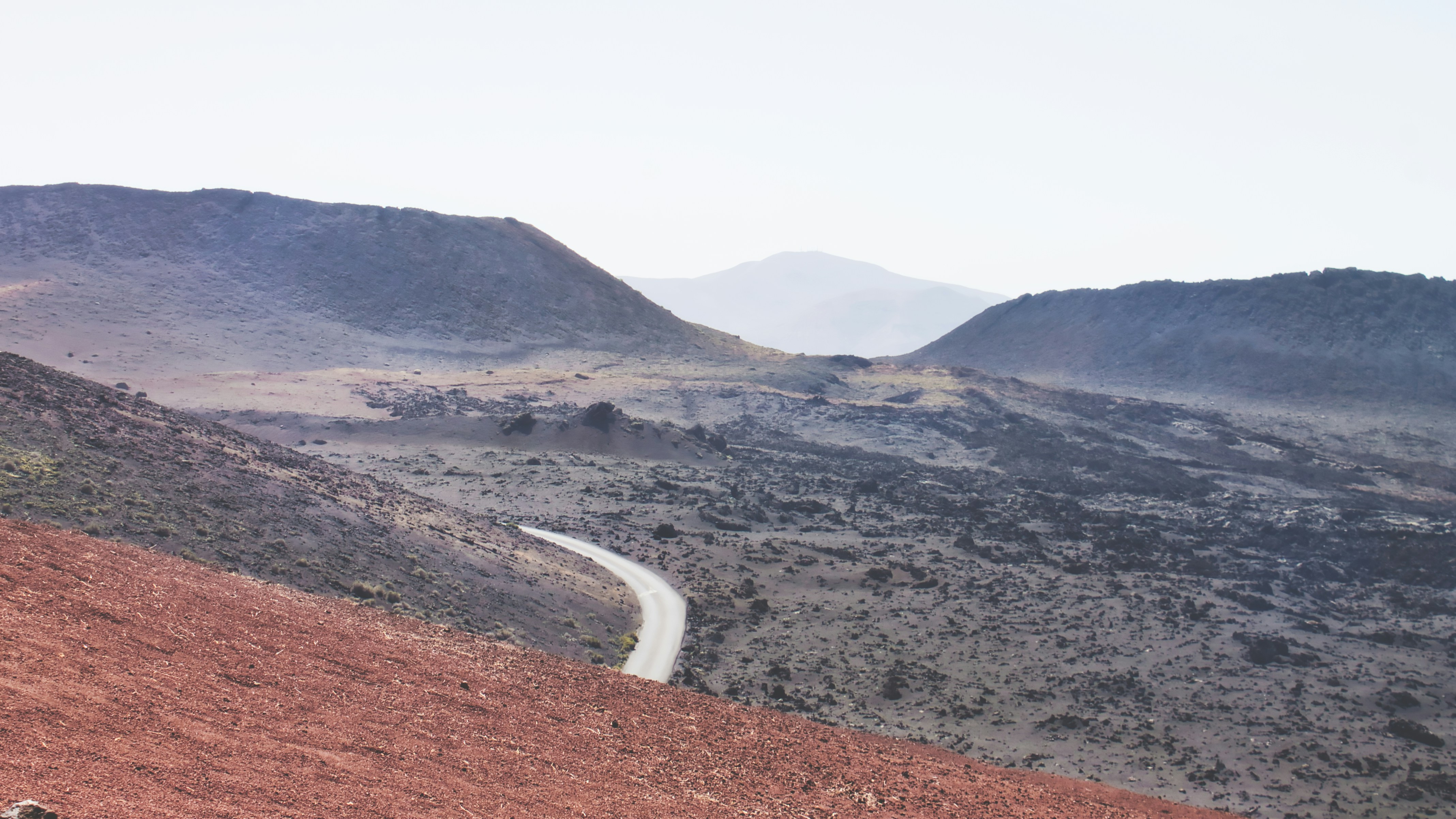 A winding road meanders through a rugged volcanic terrain, characterized by reddish earth and distant mountains. The scene captures the stark beauty of the landscape.