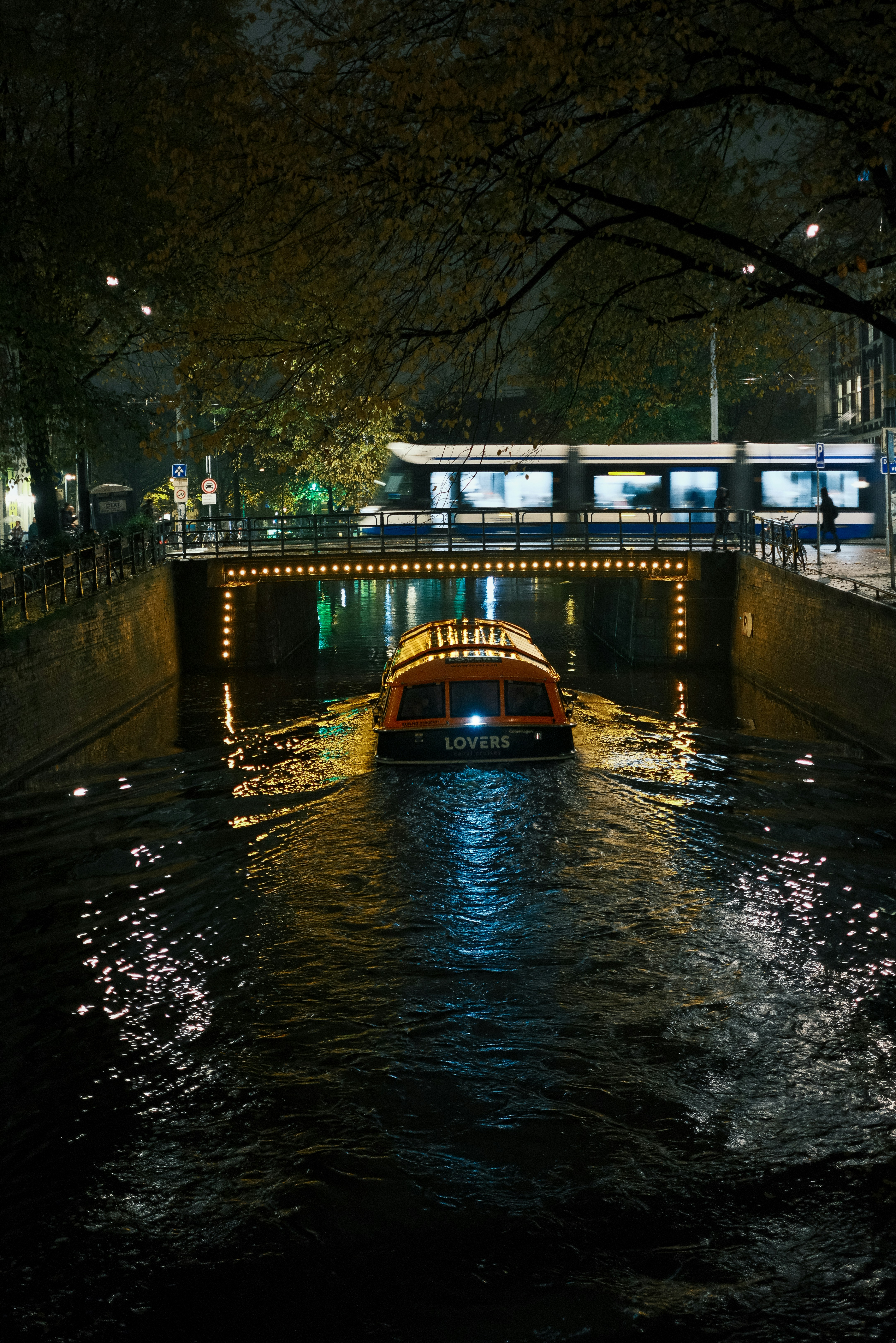 A boat named 'LOVERS' glides through a dimly lit canal under a bridge, while a tram passes overhead, creating a dynamic urban scene.