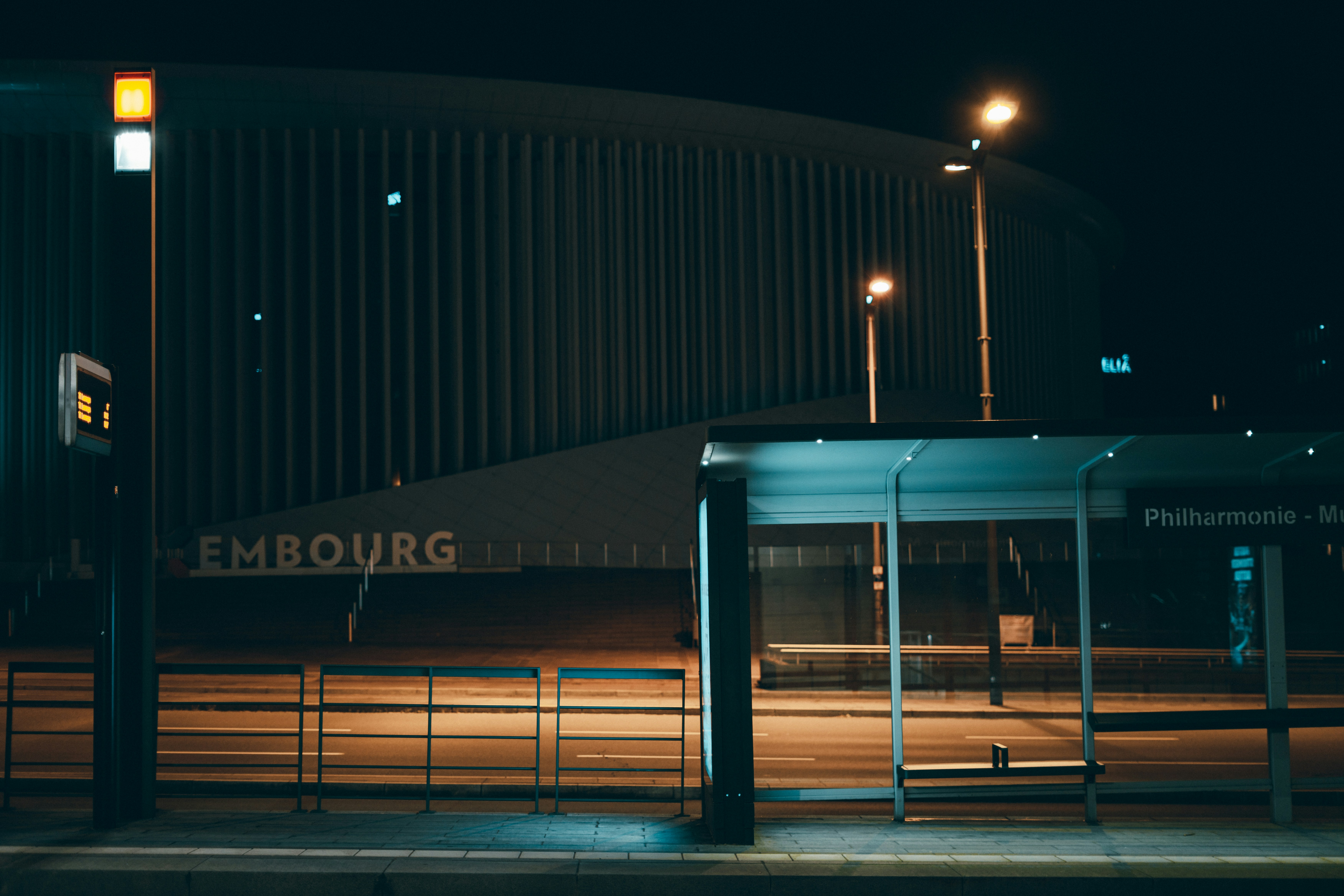 Bus stop illuminated by streetlights, with a modern architectural backdrop featuring curved lines. Nighttime urban scene showcasing the interplay of light and architecture.