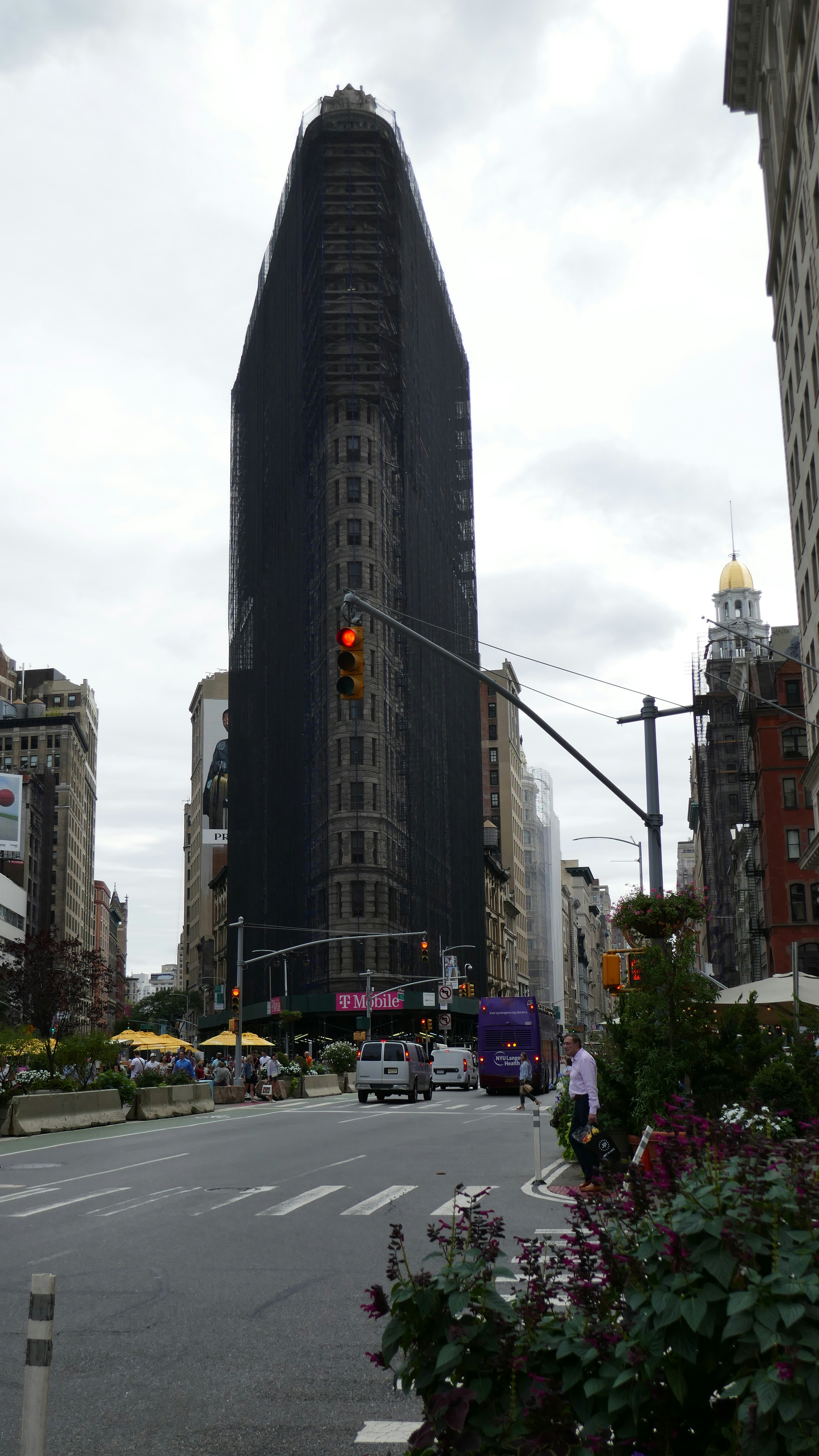 Construction-draped Flatiron-like tower dominates a busy Broadway street with pedestrians, vehicles, and a T-Mobile storefront in the background.