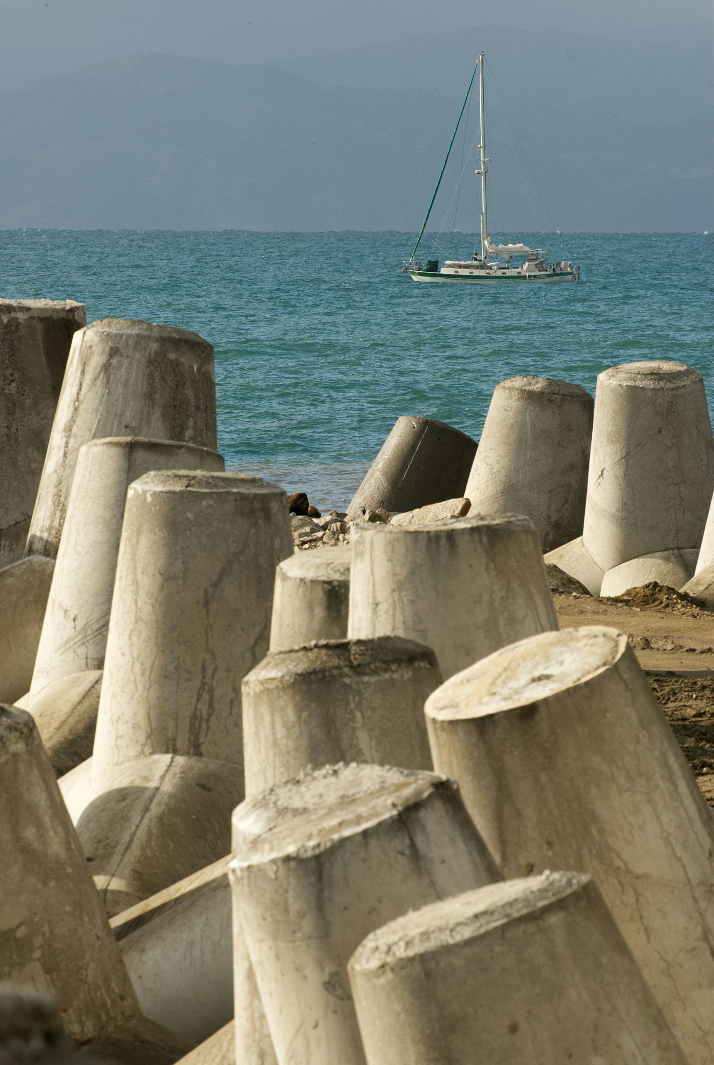 Concrete breakwaters jutting into the sea with a lone sailboat in the background against a serene blue backdrop.