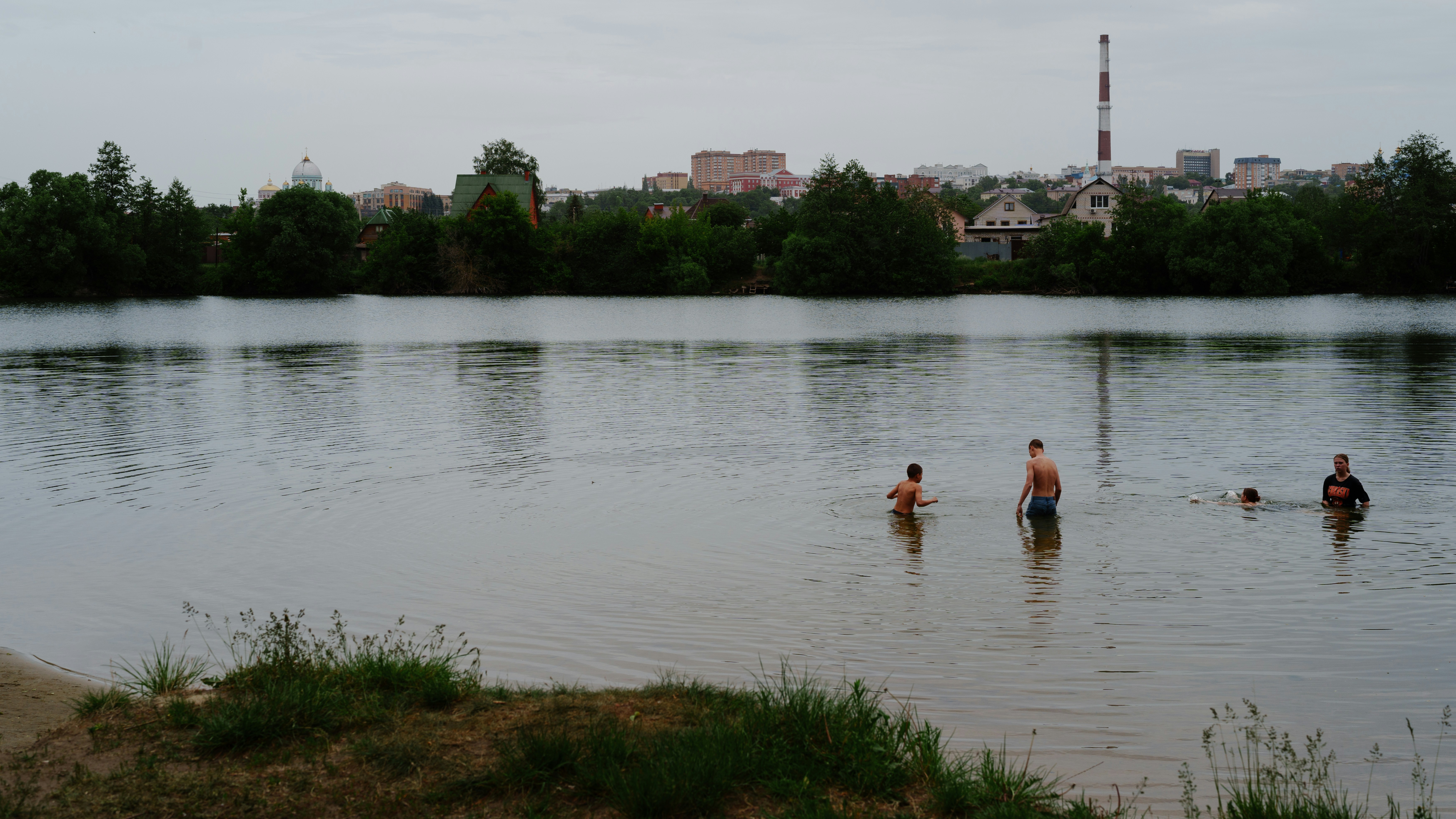 Group of people enjoying a leisurely swim in a calm river, with a backdrop of a city skyline under overcast skies.