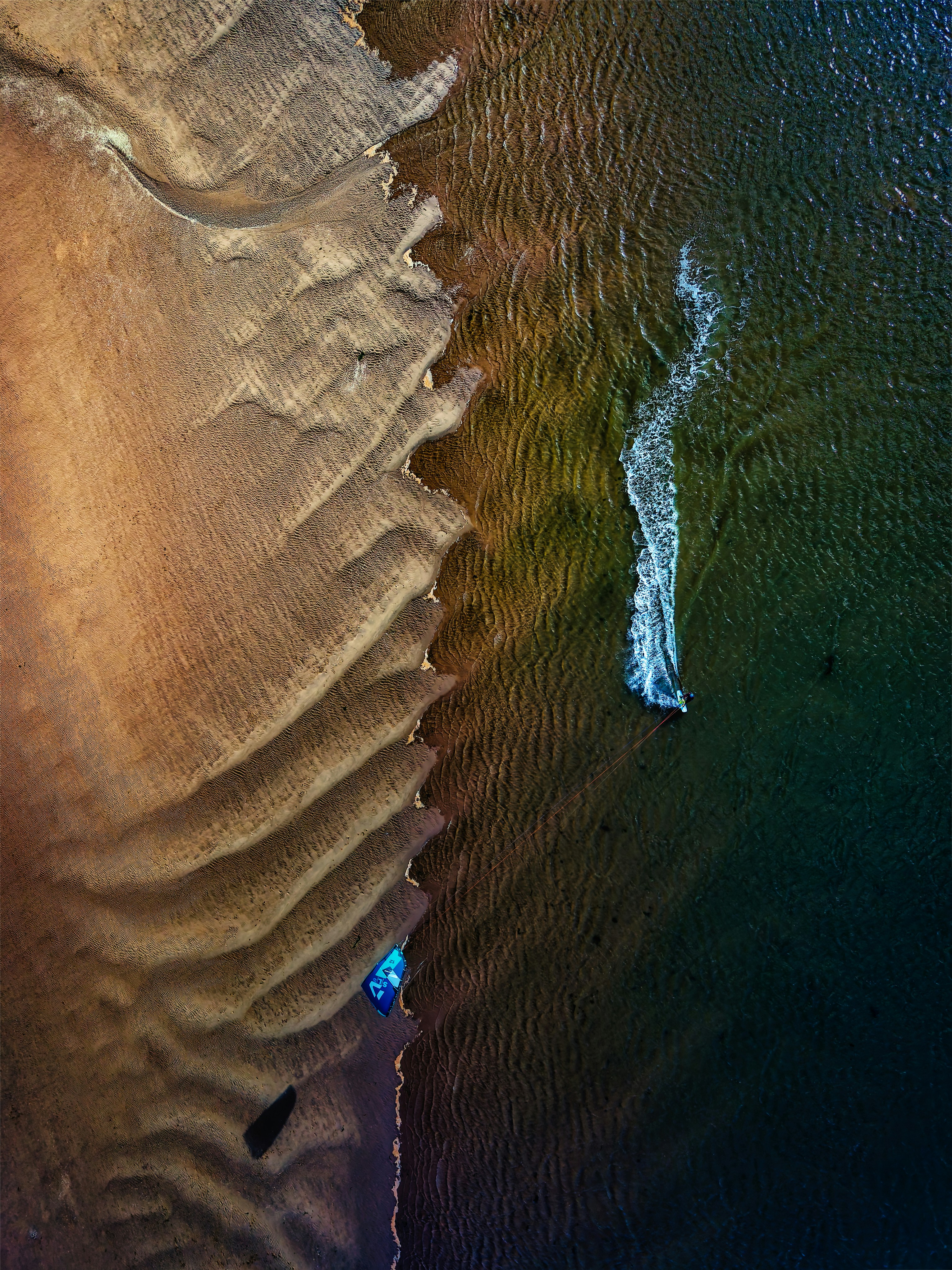Aerial view of a speedboat gliding through the water, contrasting with the textured sandy shoreline. The dynamic patterns of the waves and sand create an intriguing visual narrative.