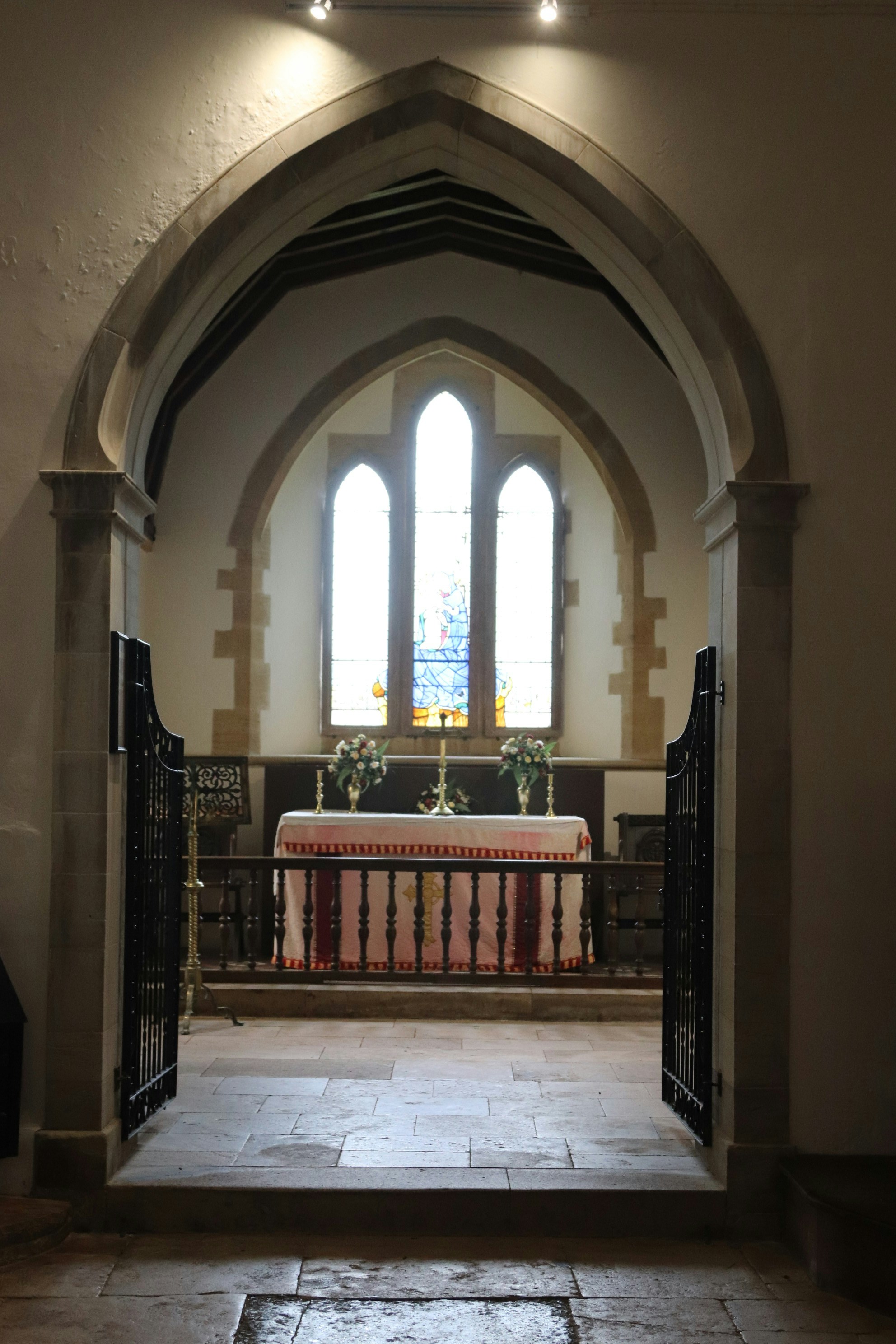 Intricate stone archway framing a serene altar adorned with floral arrangements, illuminated by stained glass windows. The atmosphere evokes a sense of peace.