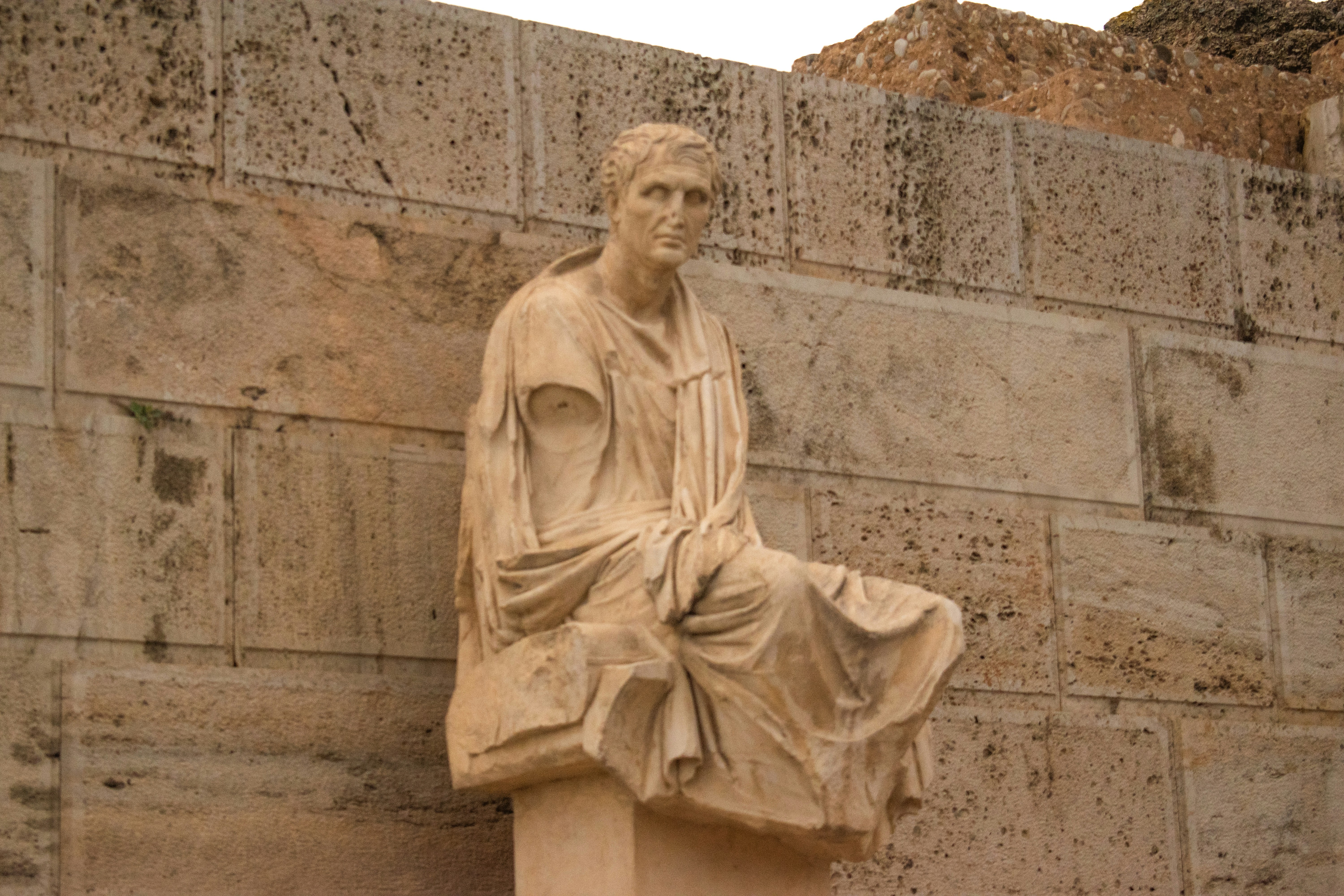 Seated marble statue of a philosopher with a contemplative expression, set against a weathered stone wall.