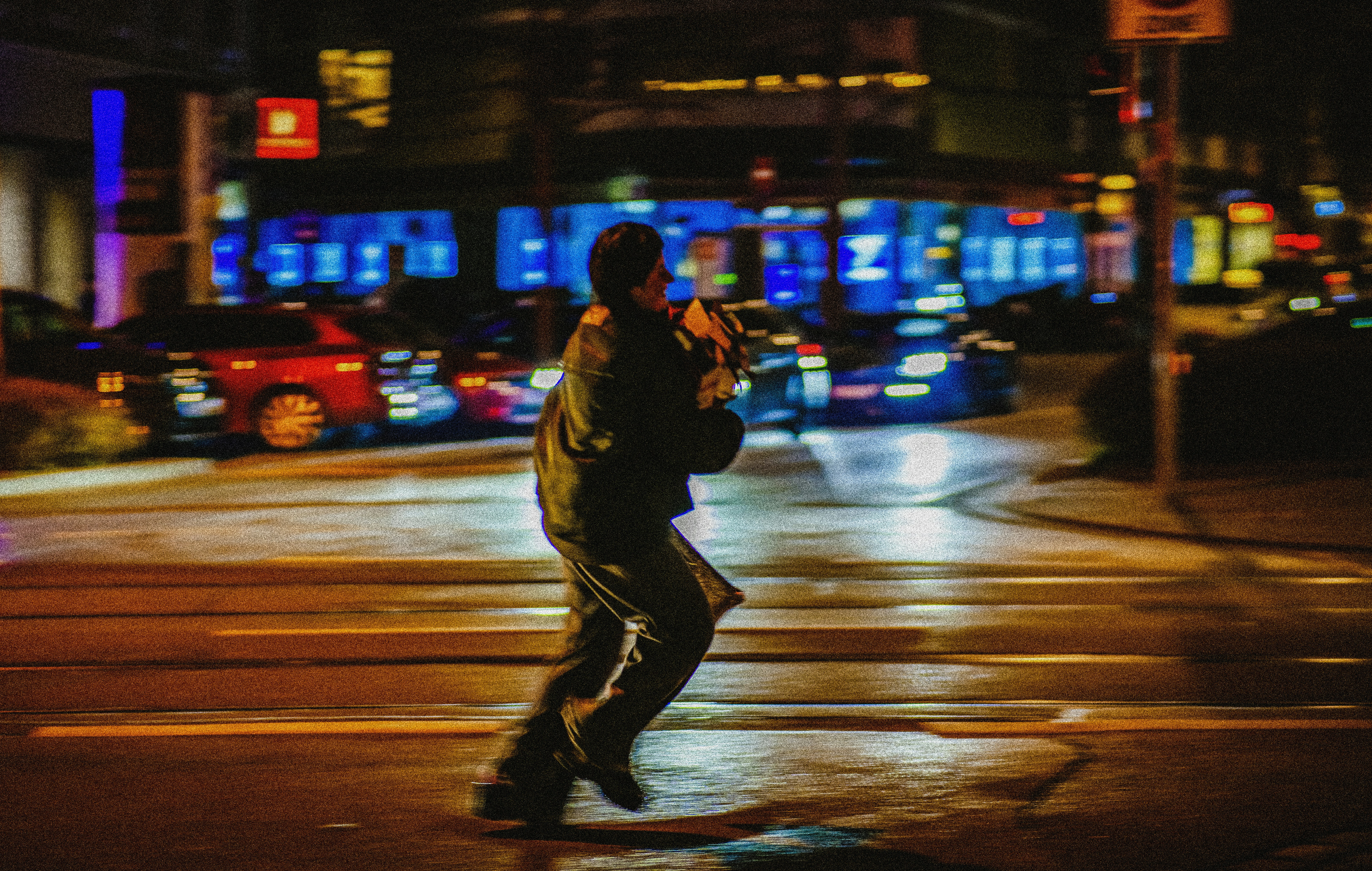 A figure hurries across a bustling urban street illuminated by vibrant neon lights, capturing the essence of nightlife in motion.