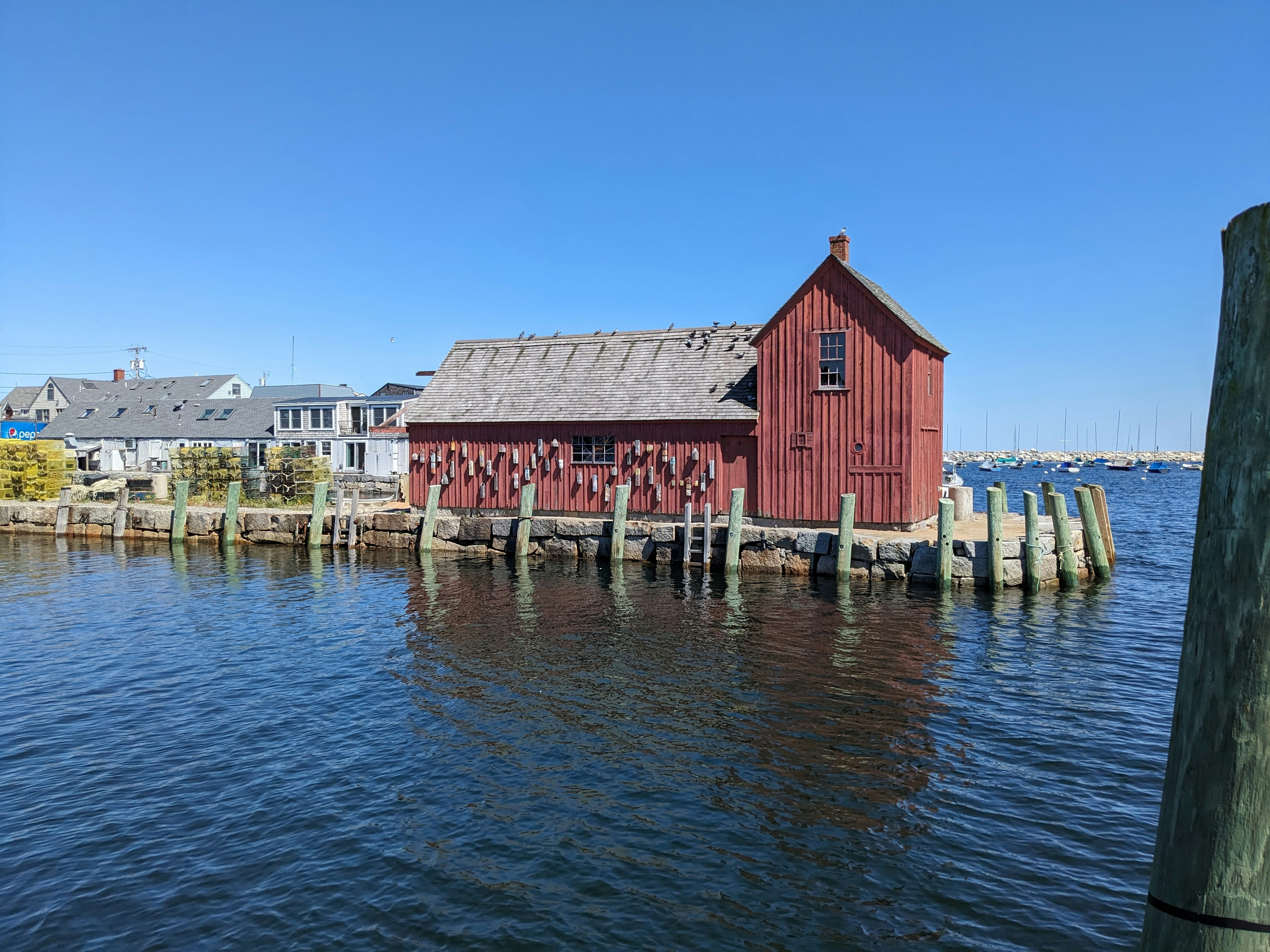 Weathered red boathouse reflecting on calm waters, surrounded by docks and boats in the distance.