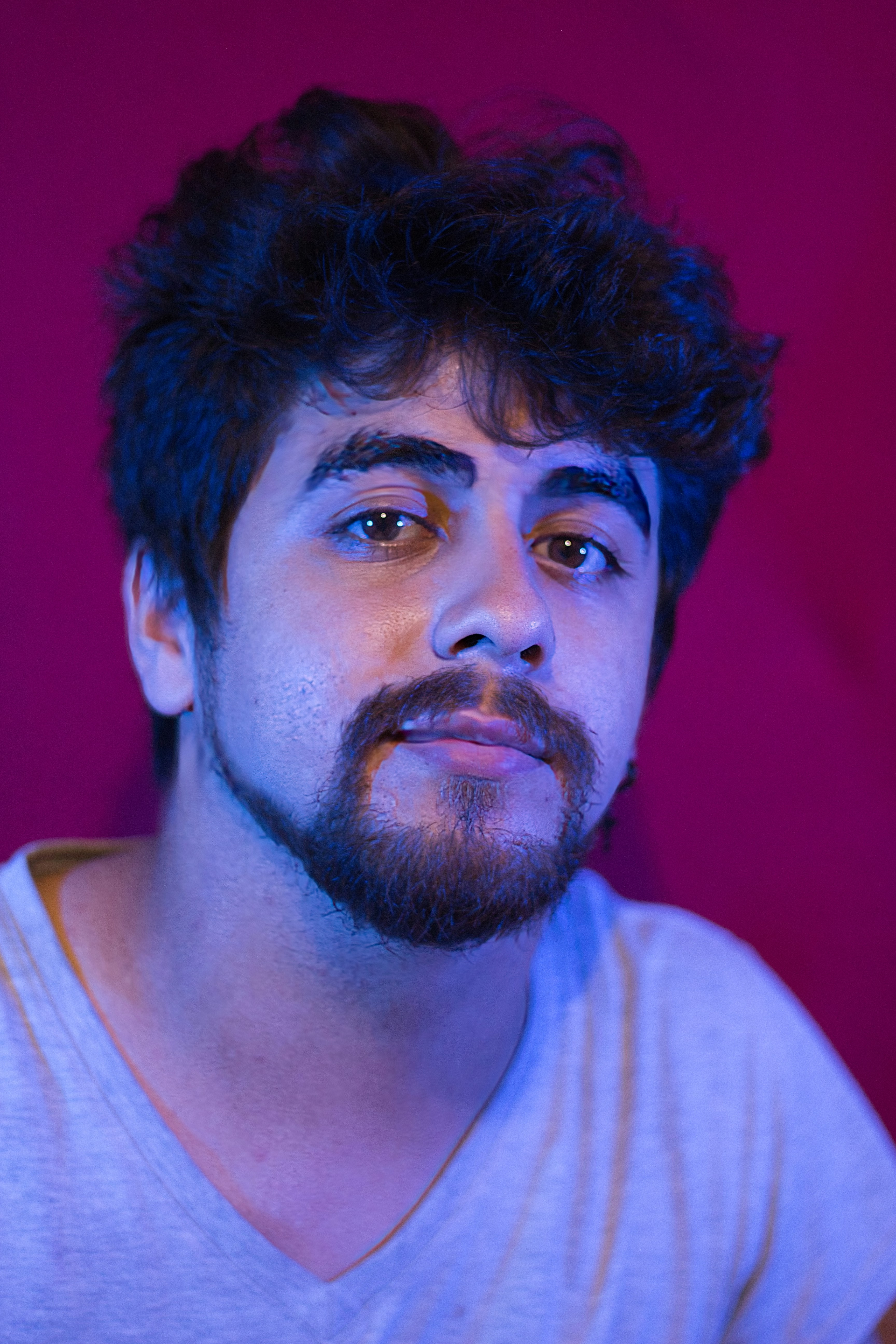 A young man with a beard and tousled hair gazes thoughtfully, illuminated by vibrant blue and purple lighting against a rich red backdrop.