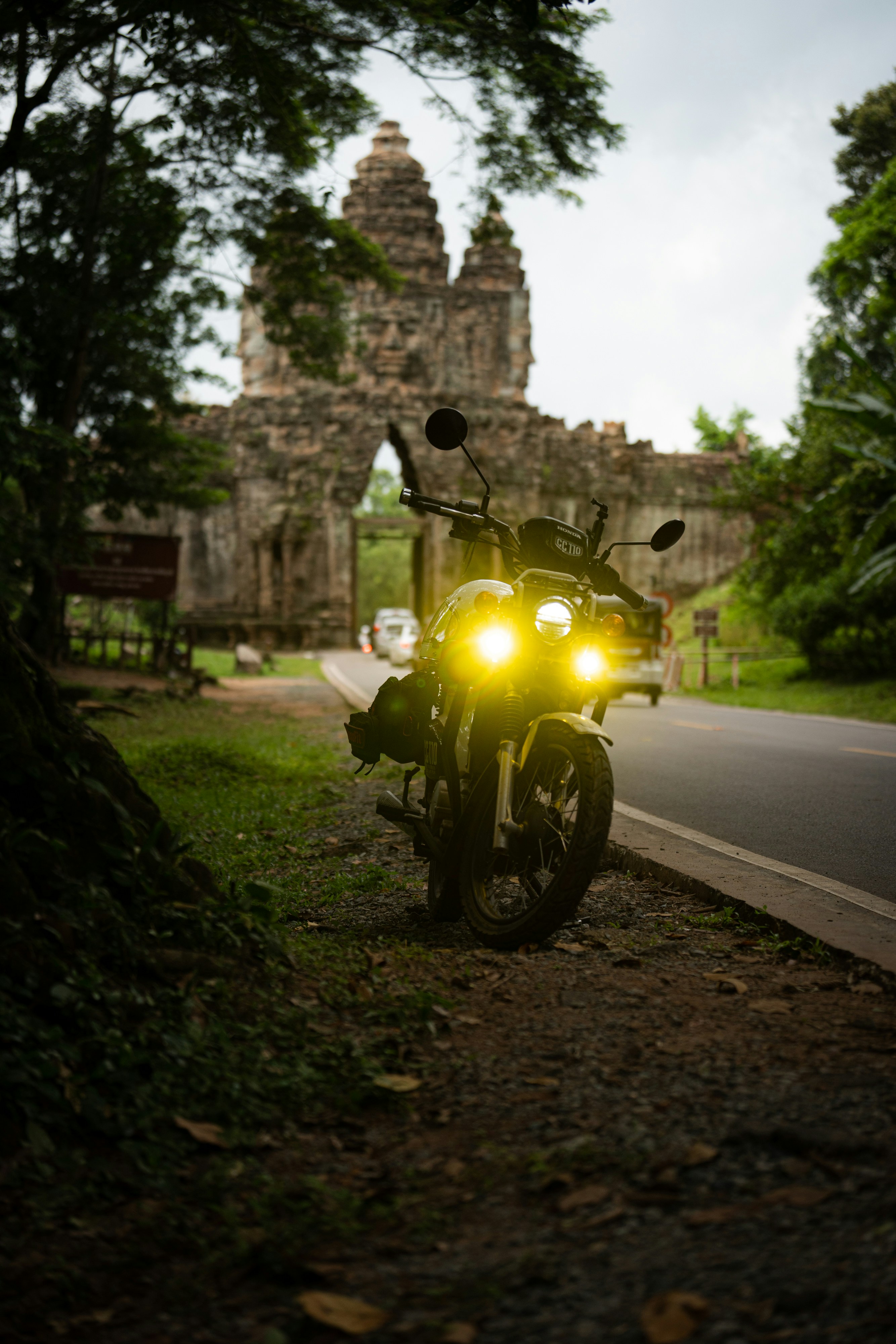 Motorcycle with illuminated headlights parked beside an ancient stone archway in a lush green setting.