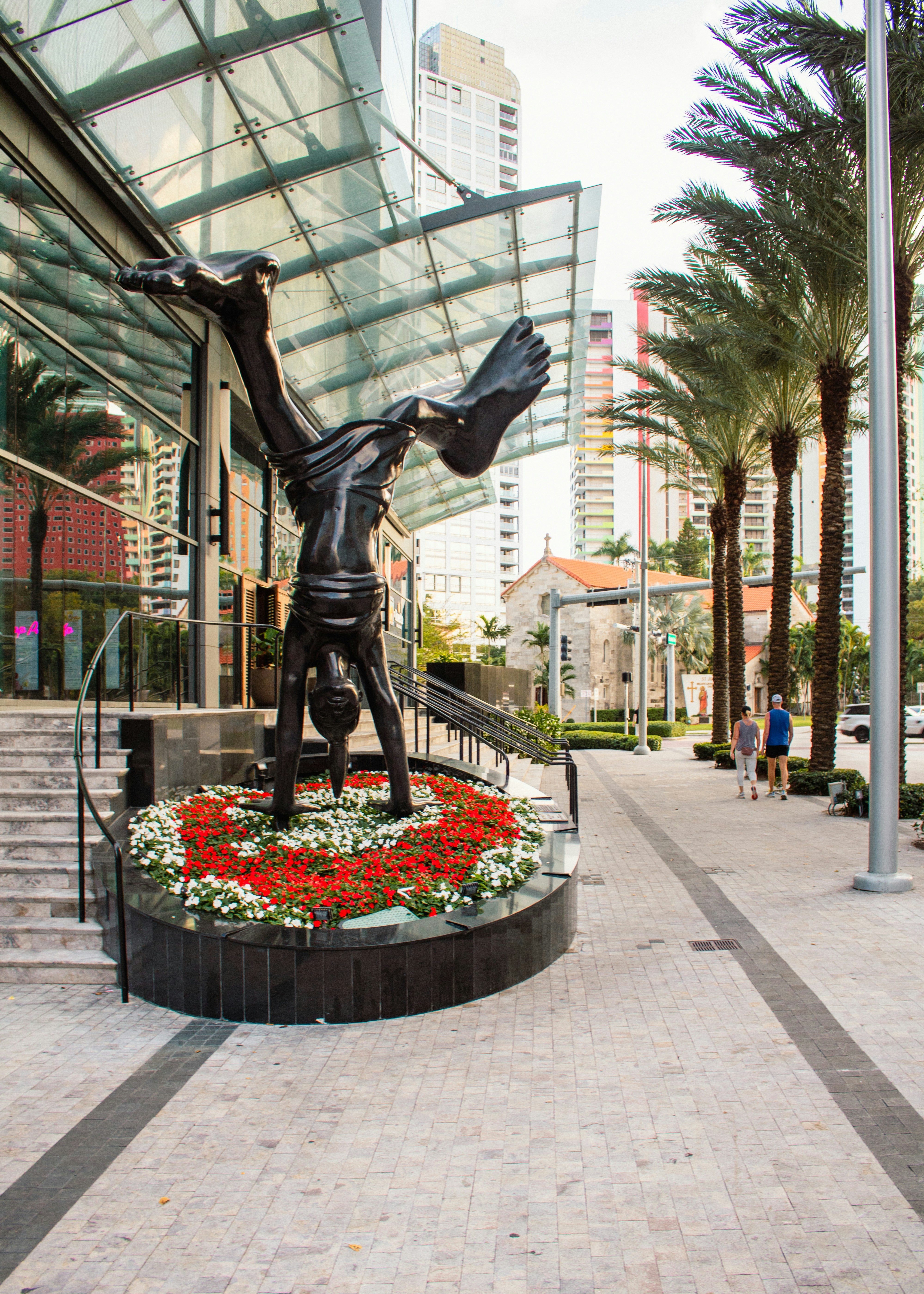 The sidewalk outside Echo in Brickell that is decorated with palm trees and a beautiful sculpture. | A statue performs a handstand in front of a building.